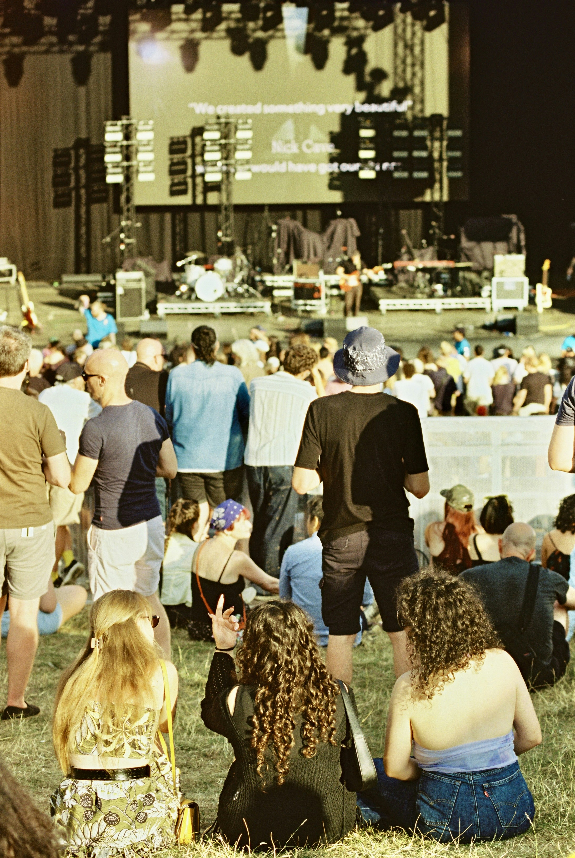 Crowd gathers in front of a stage with instruments and a large screen, capturing a live concert scene.