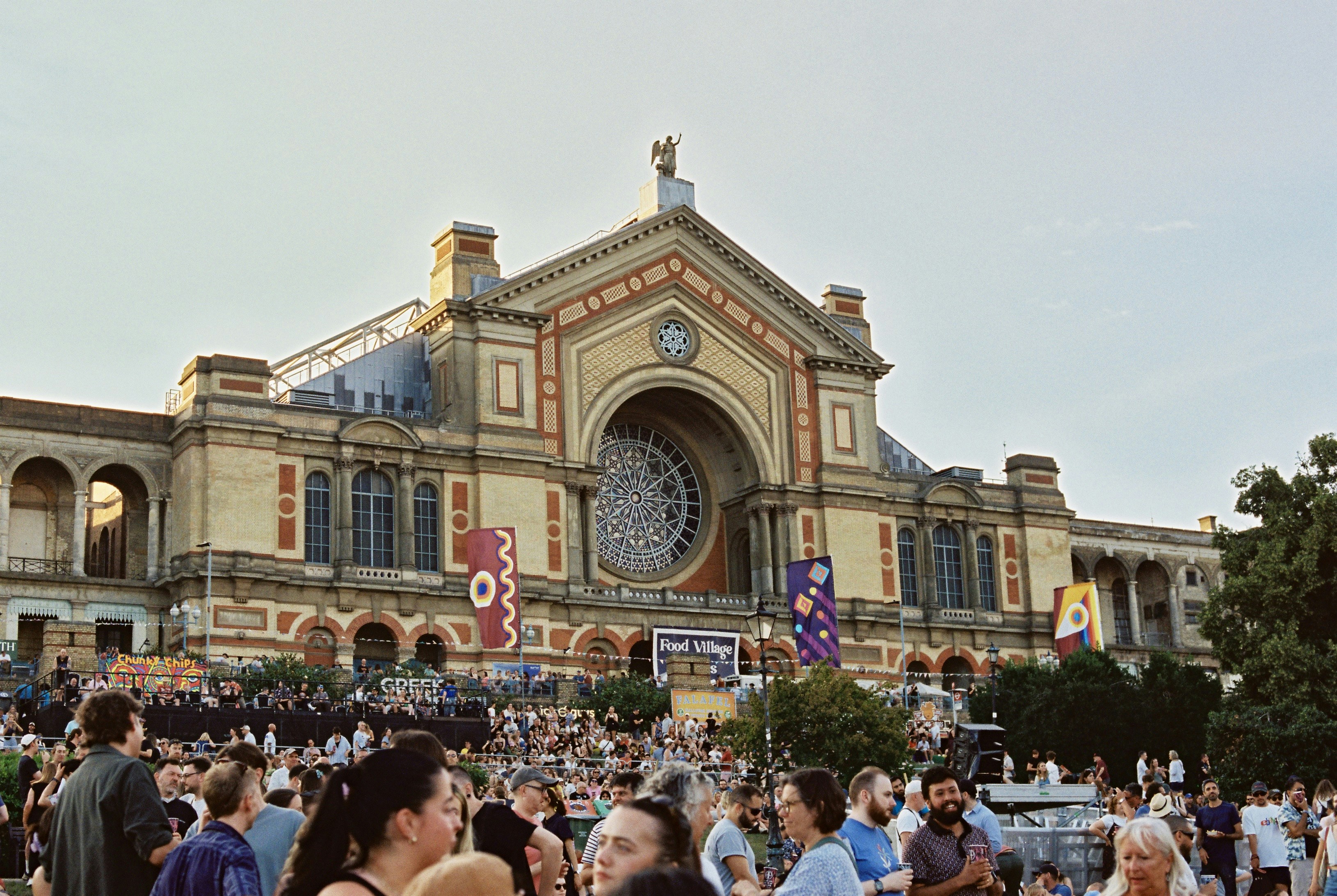 Crowd gathers in front of a historic railway station during a daytime festival, with banners and people occupying the plaza and steps. A circular stained-glass window anchors the facade.