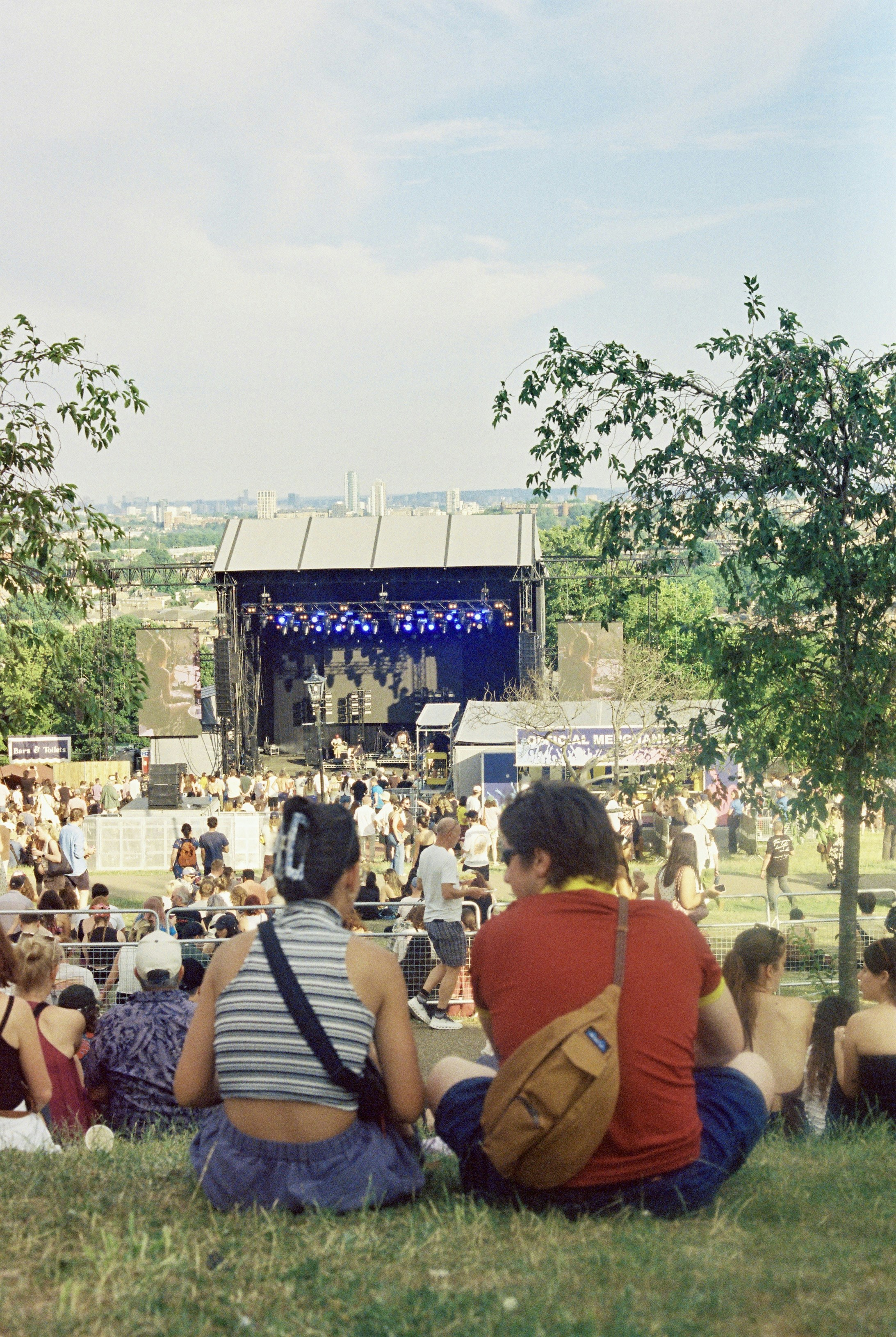 Two friends in a sunlit park festival watch a distant concert from the crest of the hill