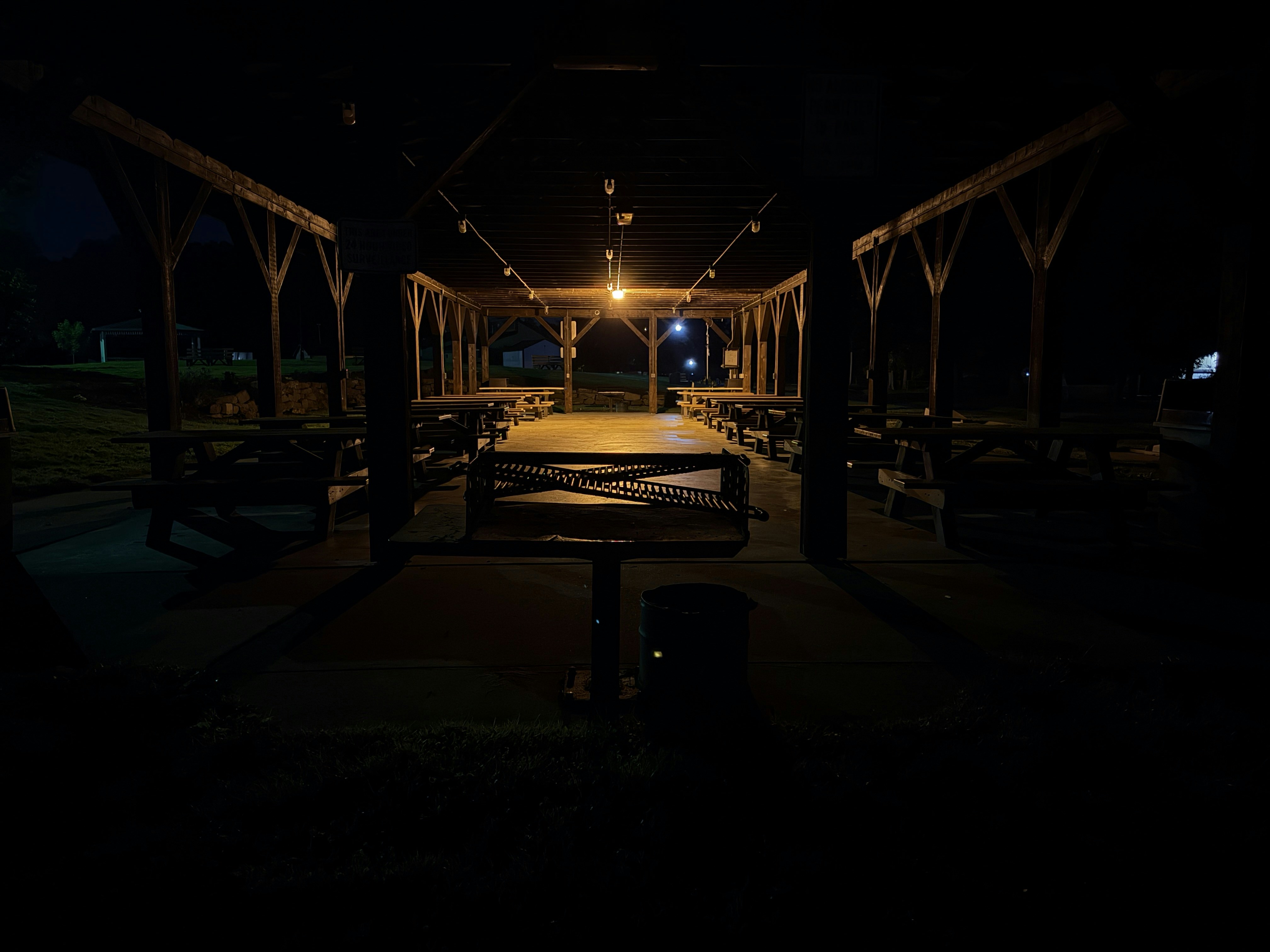 Empty park pavilion at night. A wooden park pavilion glows in warm amber light against a pitch-black night, its symmetrical timber frame and rows of empty picnic tables receding into darkness while a single bulb illuminates the center like a lantern.