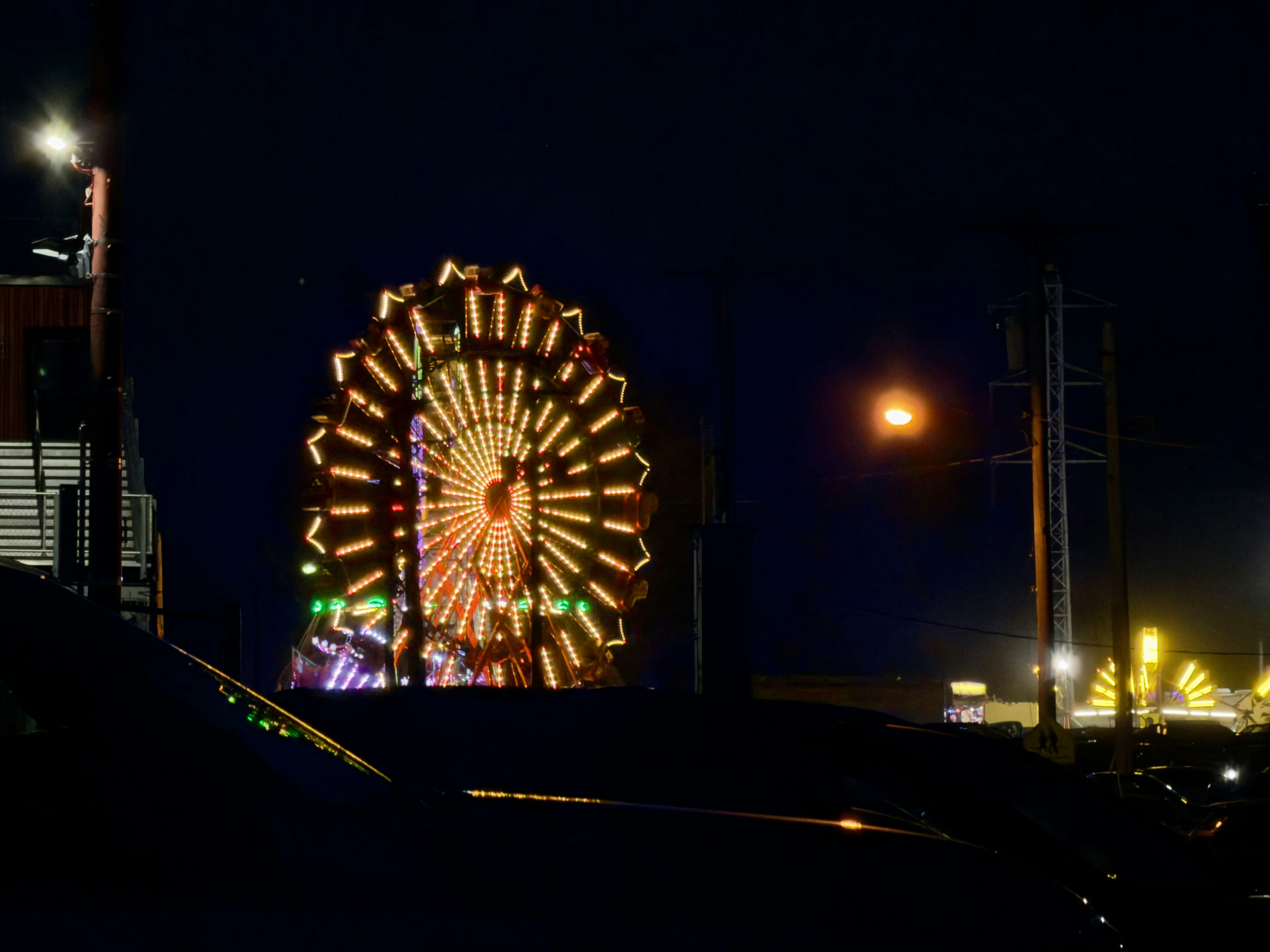 Brightly lit Ferris wheel blooms with warm neon against a dark urban nightscape, with silhouettes of vehicles and streetlights in the foreground.