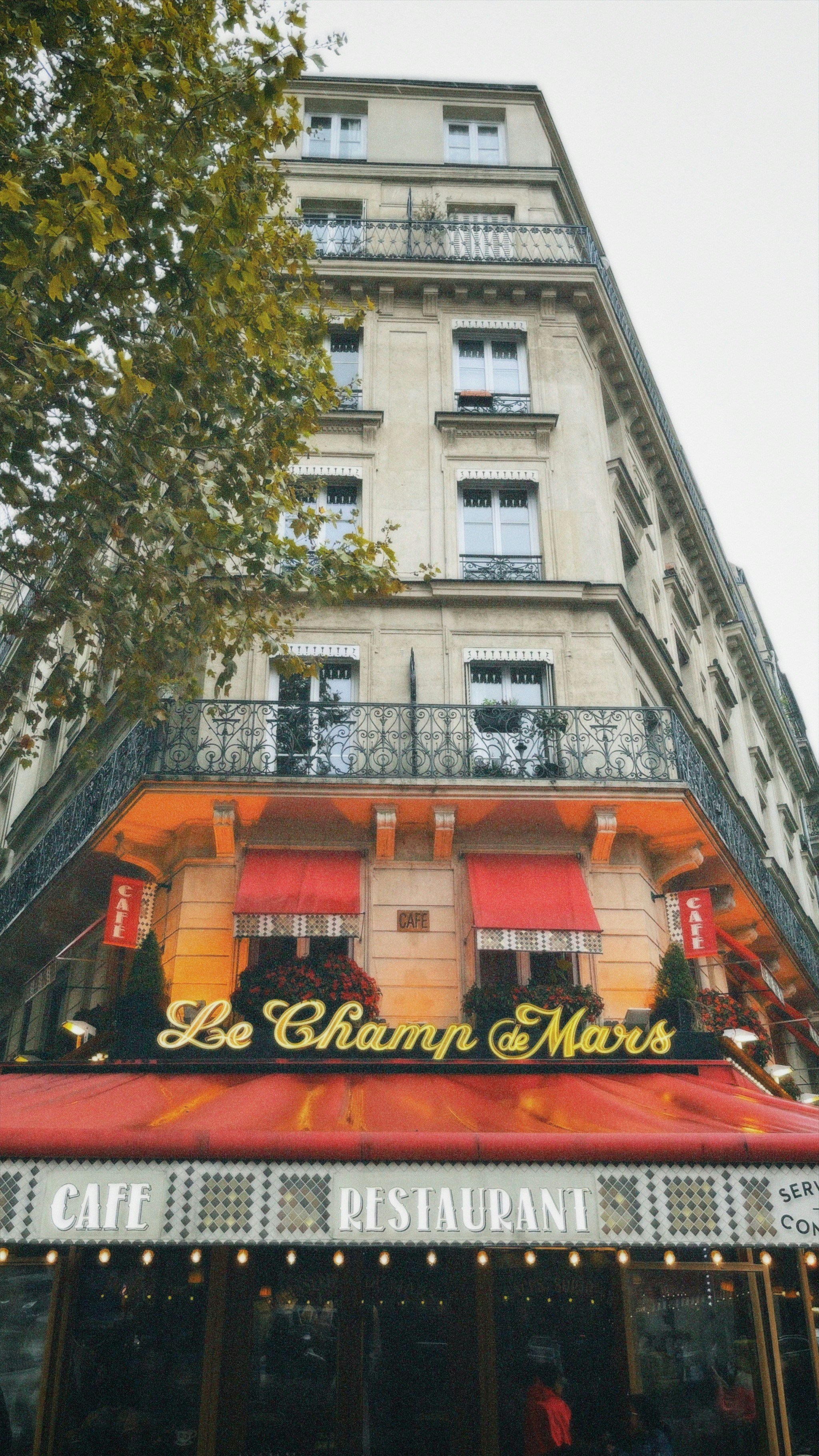 Tall Parisian building with a cafe and red awnings anchors the frame; warm neon signage beneath a row of balconies contrasts with the stone facade.
