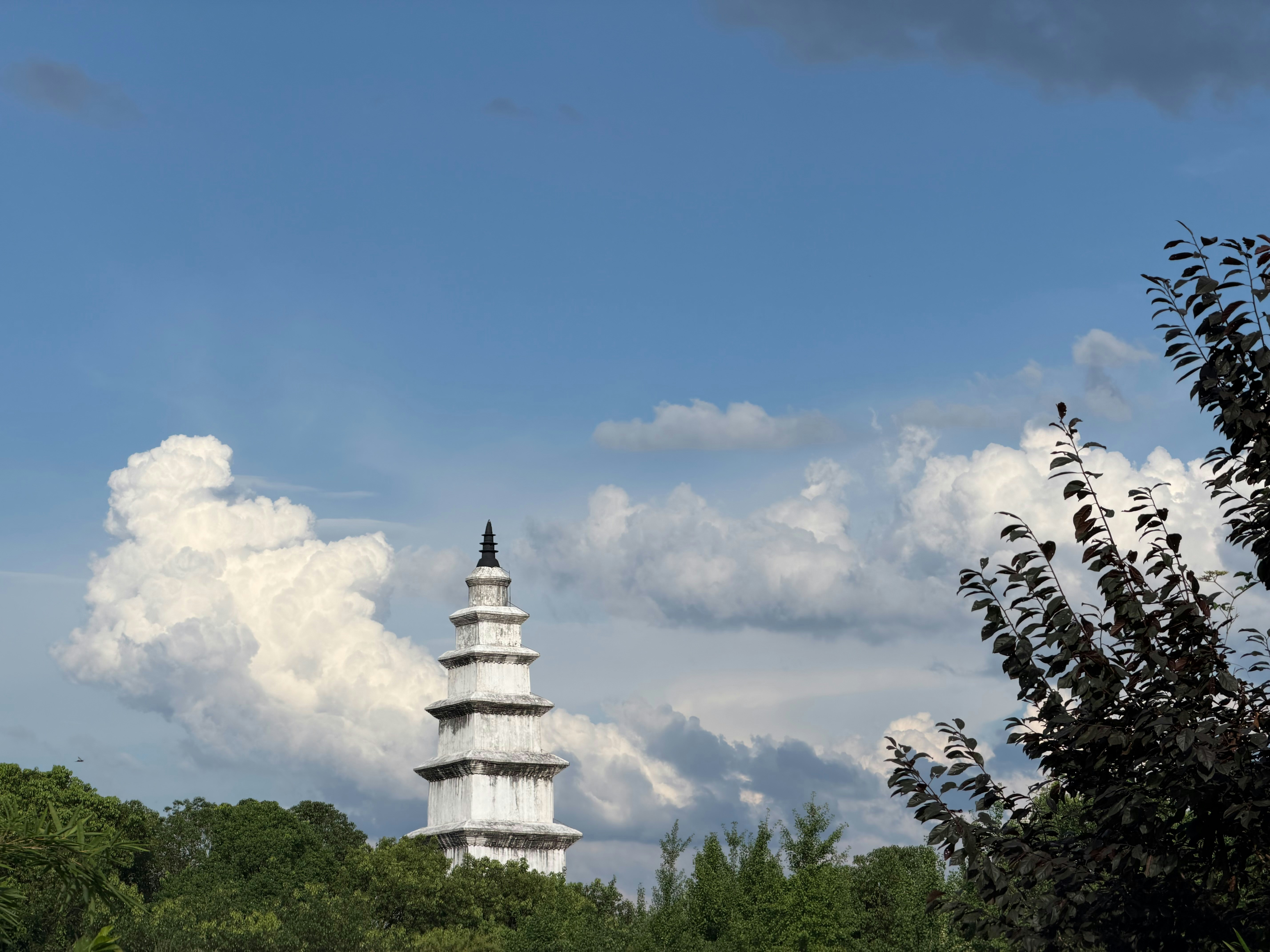A white multi-tiered pagoda rises above a lush treeline under a blue, cloud-filled sky. The scene emphasizes tranquil landscape and architectural contrast within a natural setting.