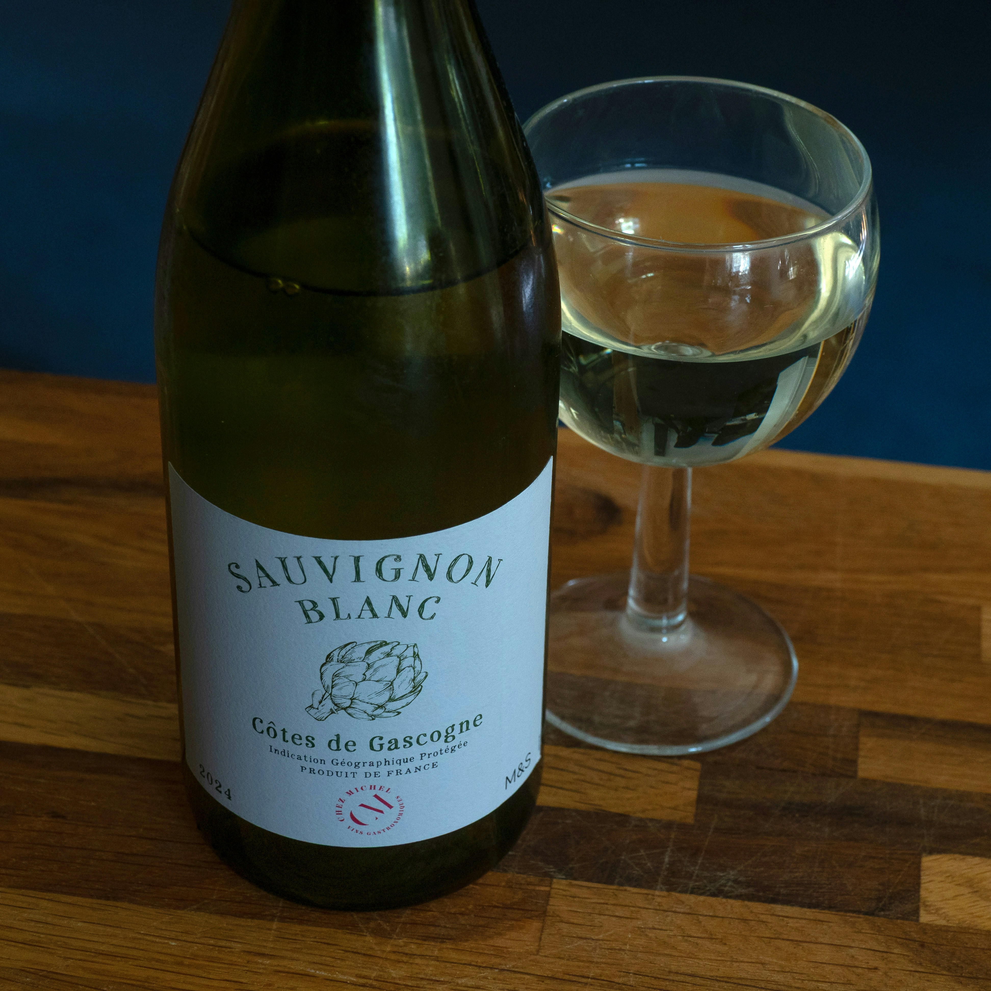A bottle of Sauvignon Blanc sits beside a glass of wine on a wooden surface against a dark blue background. The composition emphasizes the label and the glass as the tasting focal points.