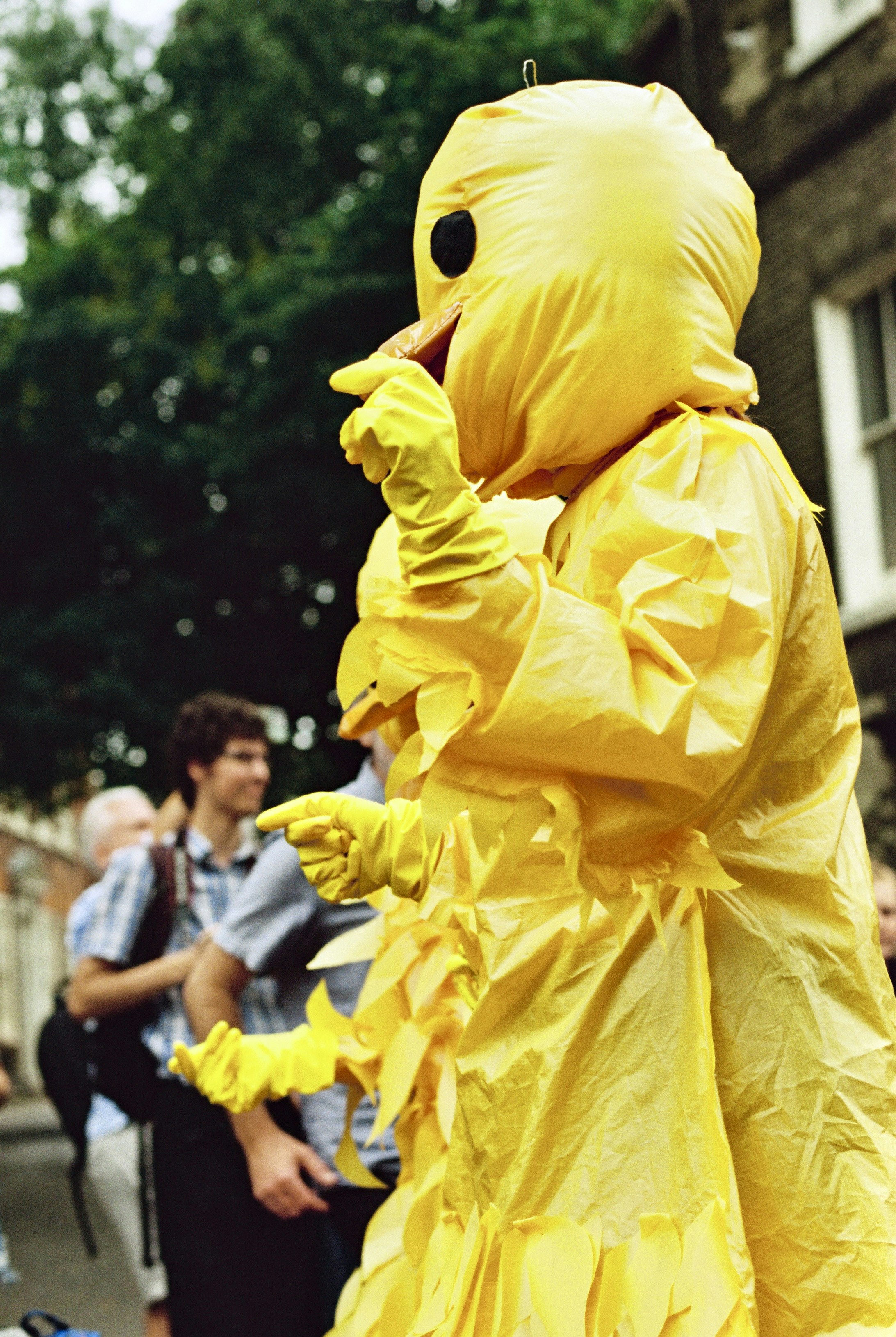 A performer in a bright yellow duck costume gestures amid onlookers at a summer street parade.