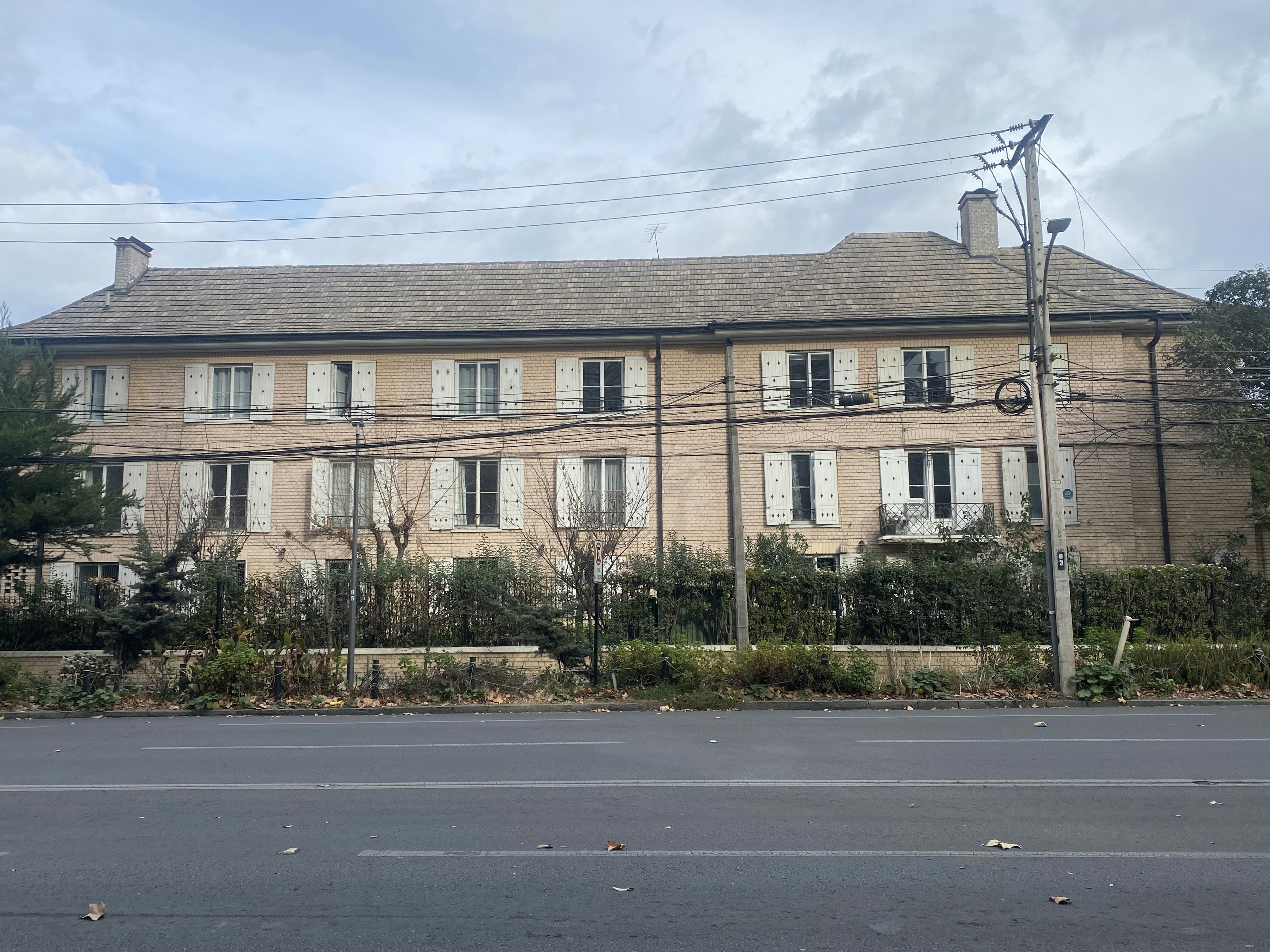 Brick apartment building with white-shuttered windows sits behind a row of trees and a fence along a wide street.