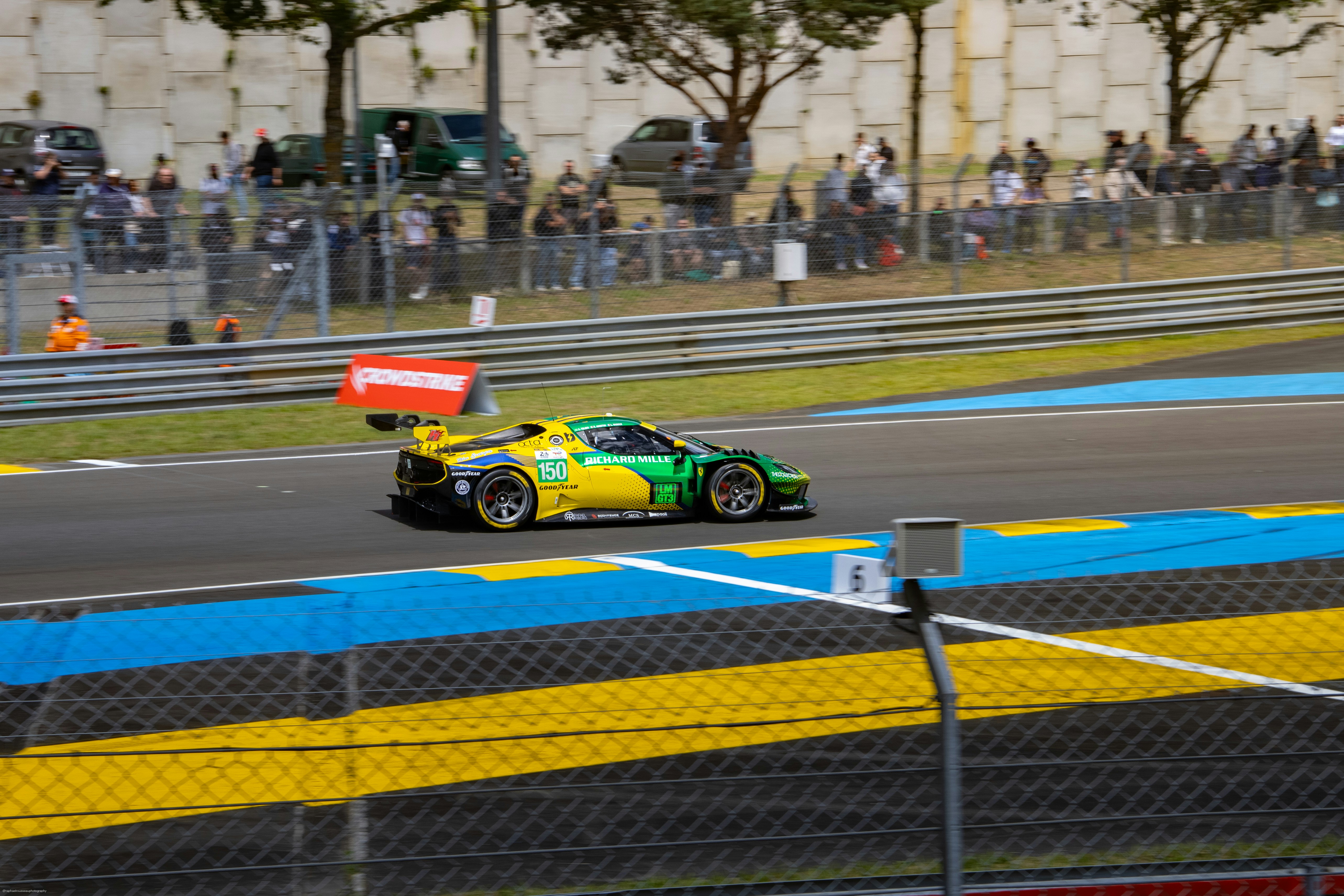 Race car speeding along track with colorful yellow and blue curbs, spectators in background; foreground fence separates track from stands.