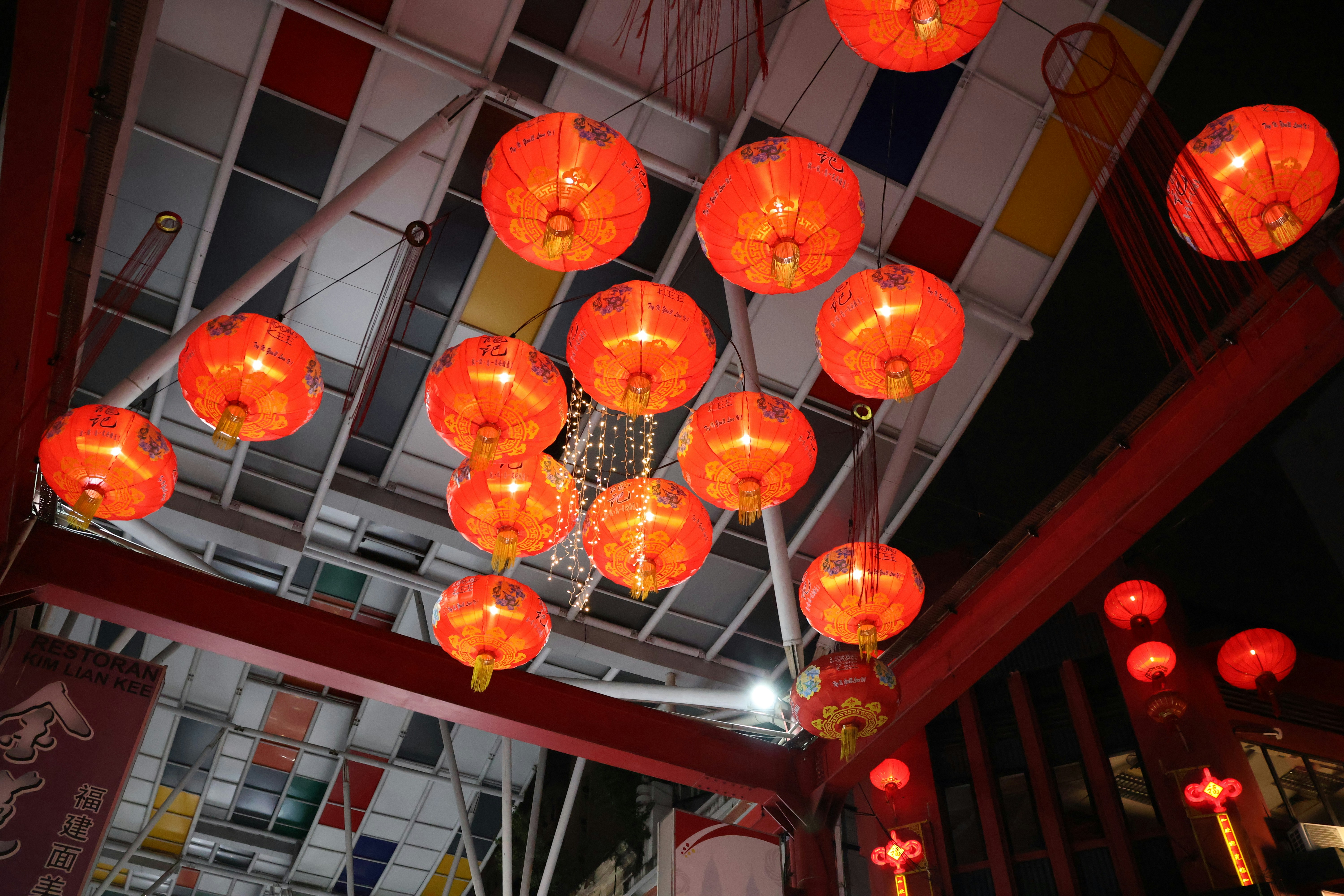 Red lanterns hang from a bright, industrial ceiling, casting a warm glow across a contemporary indoor space with geometric ceiling panels and festive decor.