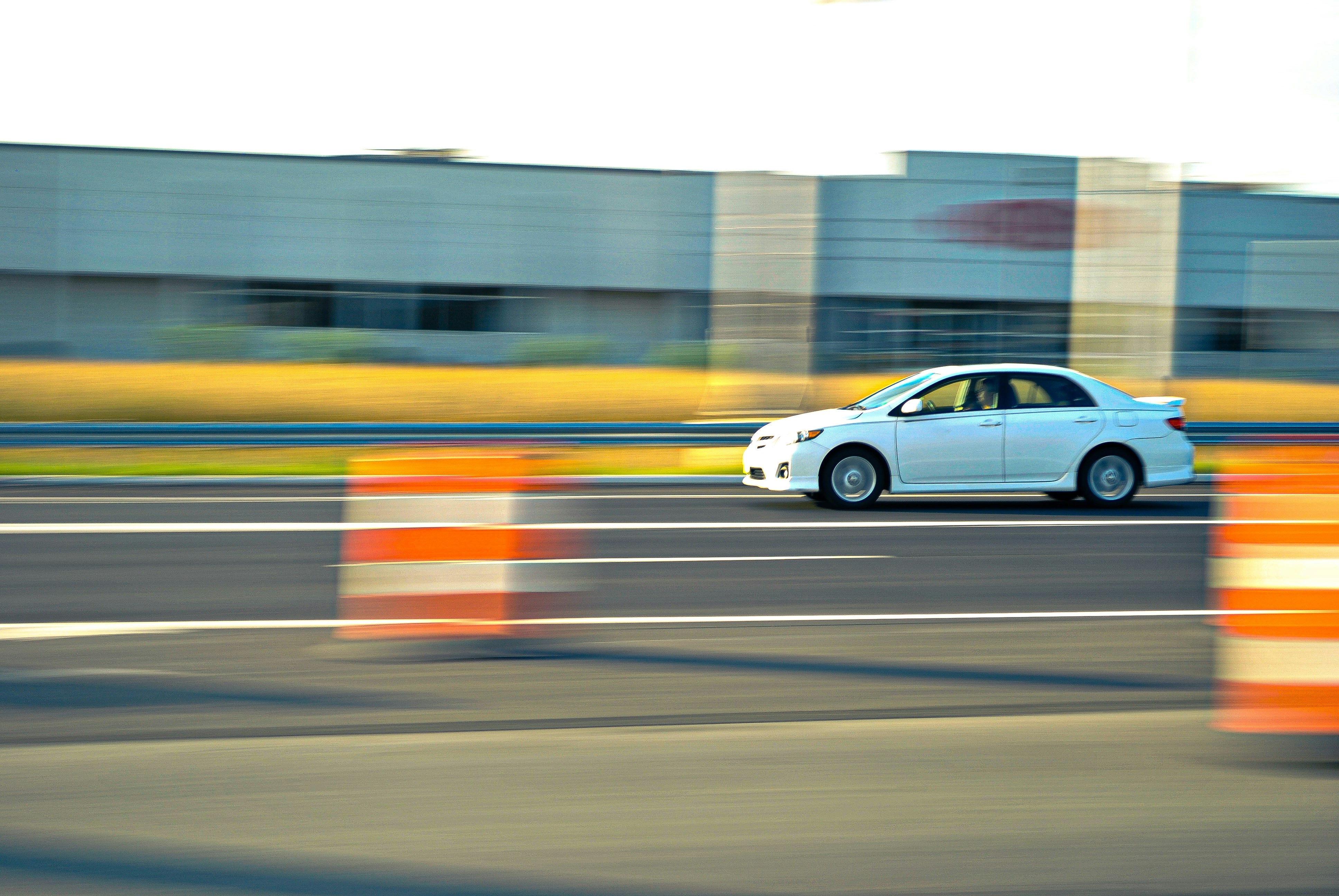 White sedan speeds along a highway with orange construction barriers in the foreground and a blurred industrial backdrop.