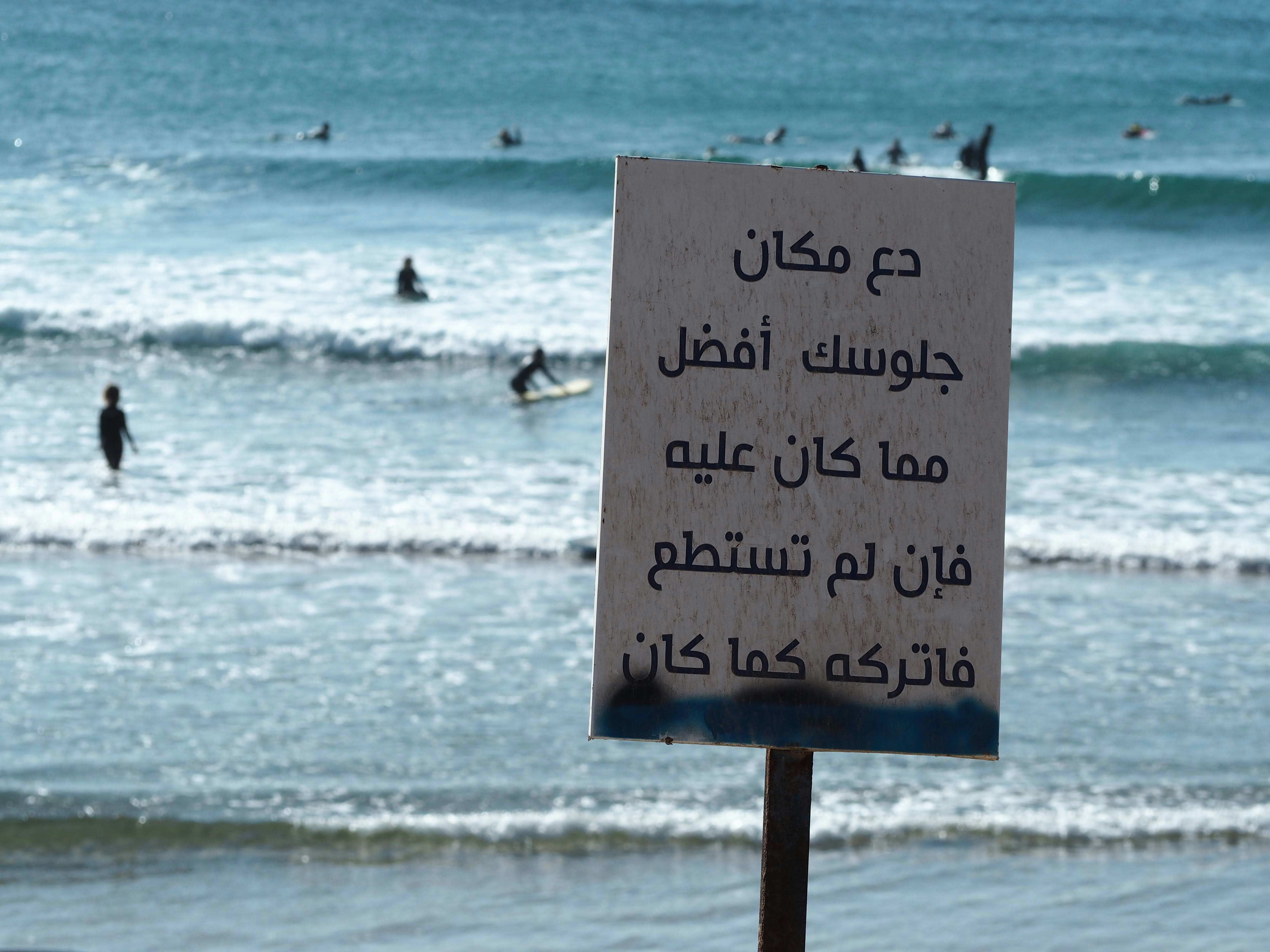 Signboard with Arabic text stands on a post near a surf-filled shoreline, water and surfers visible in the background.