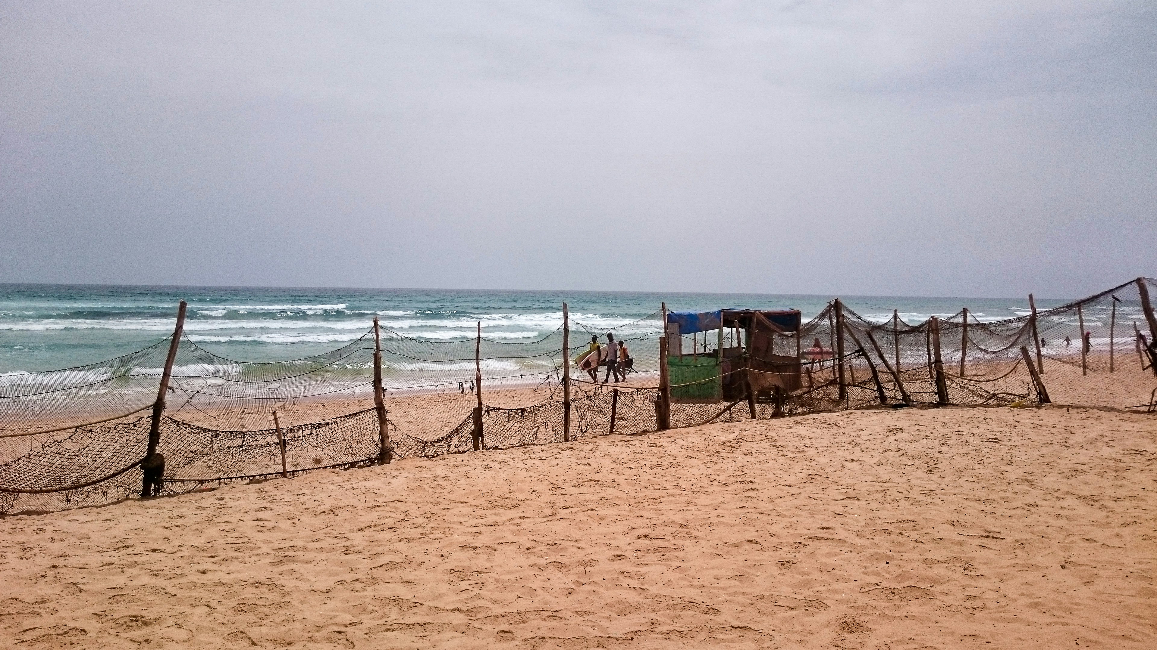 Wide beach scene with a sandy foreground, fencing and posts along the shore, and small figures near the water under a cloudy sky.