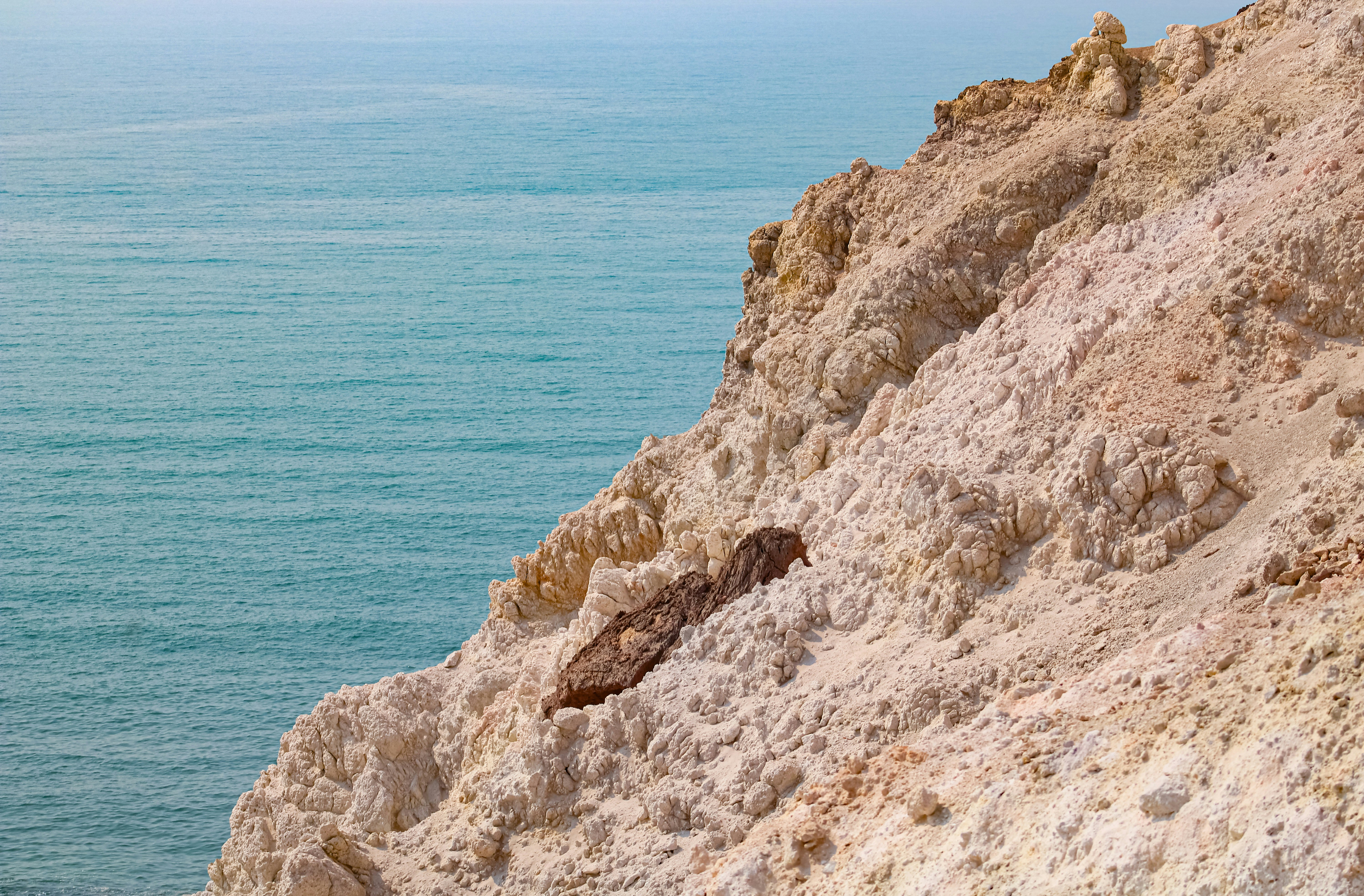 Prominent coastal cliff face rises from the sea, showing textured rock in warm tones against calm blue water; the scene emphasizes rugged geology and coastal geology without human presence. The image captures a natural landscape.