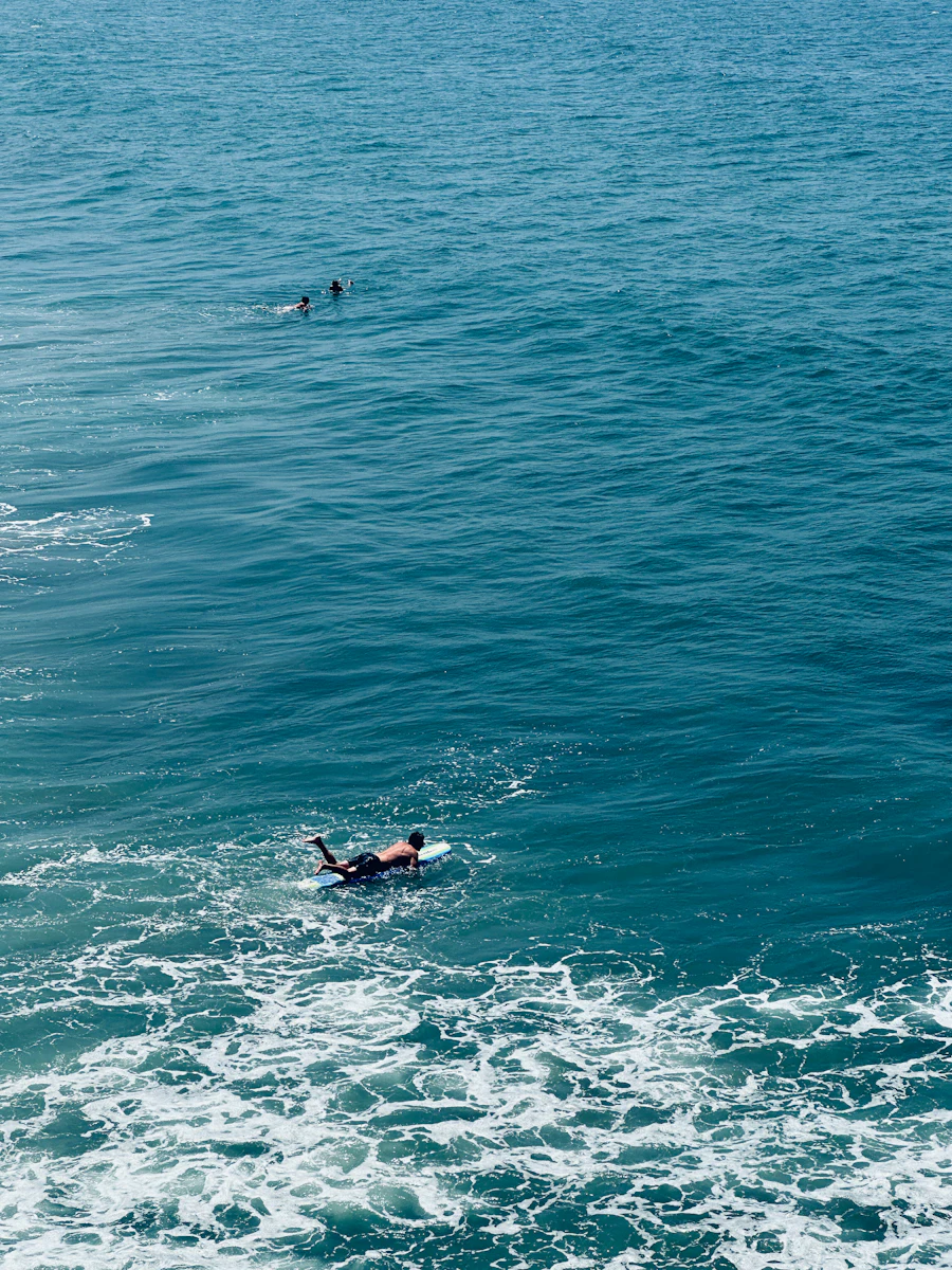 Surfer glides on turquoise sea near frothy white surf, with a distant couple visible further out in the blue open water.