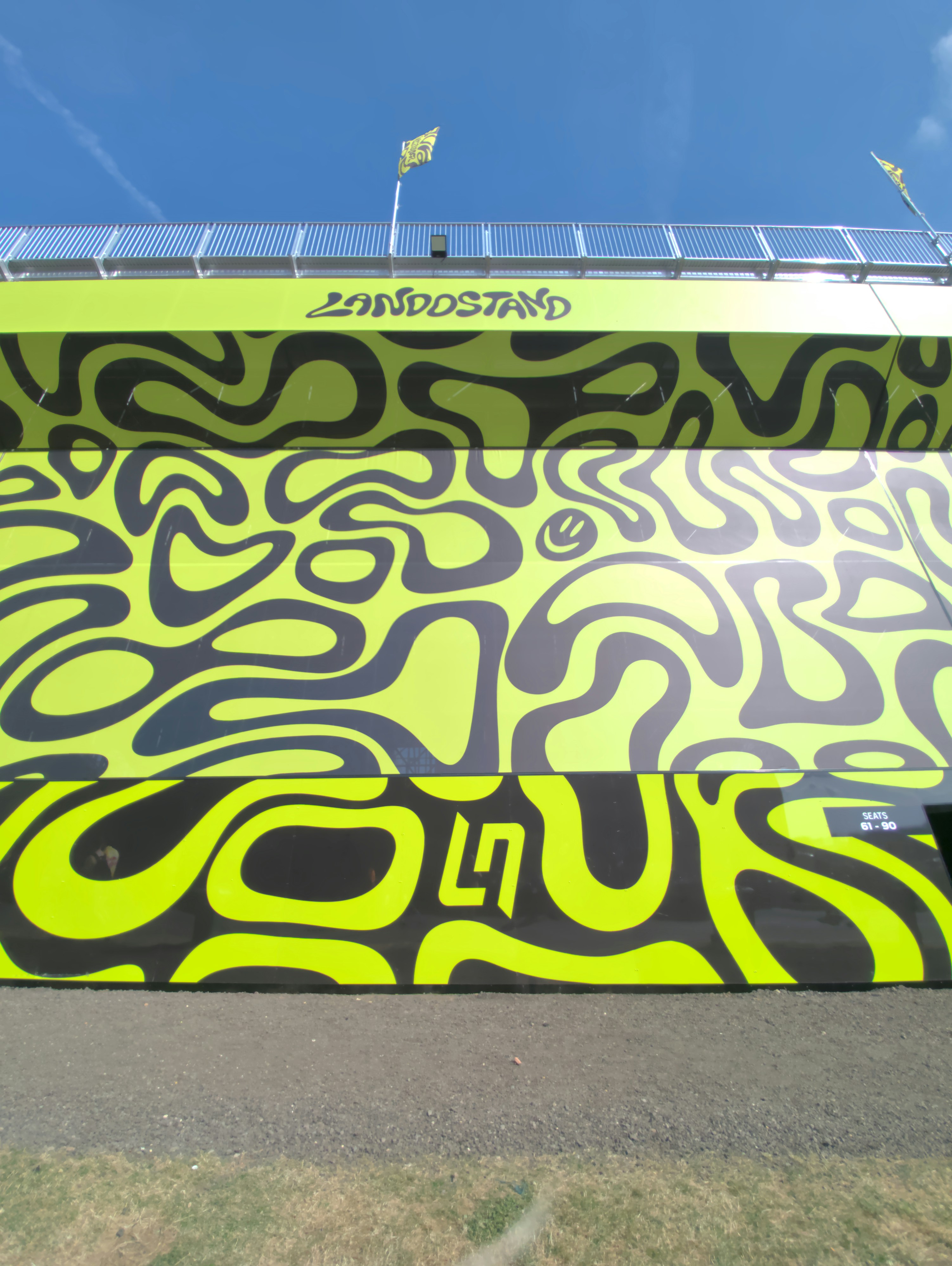 Bright lime and black abstract mural on a transport side, with blue sky above and a gray ground at the bottom; bold patterns fill the frame and a logo-like text runs along the top edge. This is a large-scale painted surface, likely a bus or trailer.