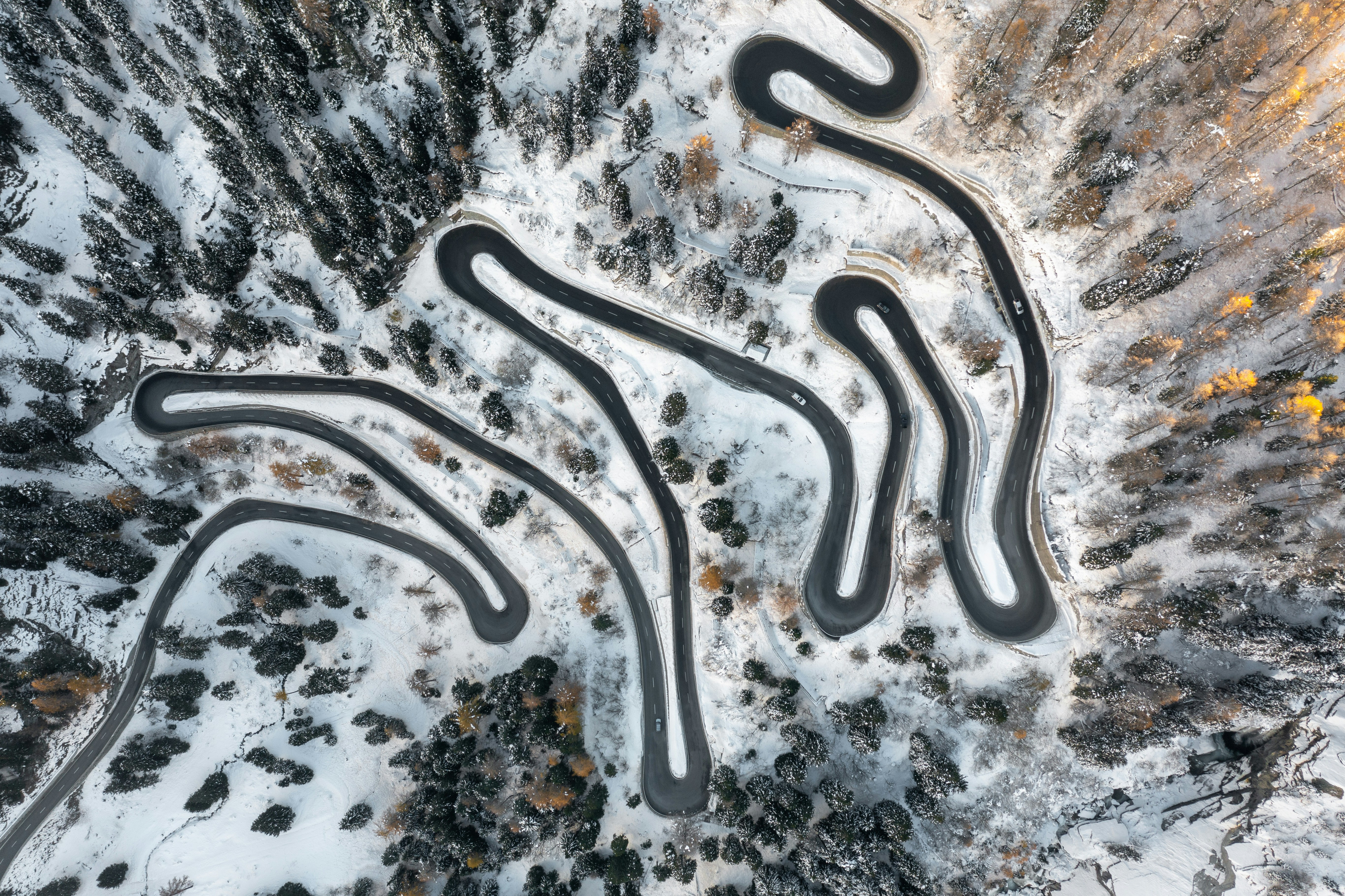 Winding mountain road snakes through a snow-dusted forest, captured from above to emphasize the pattern and seasonal contrast.