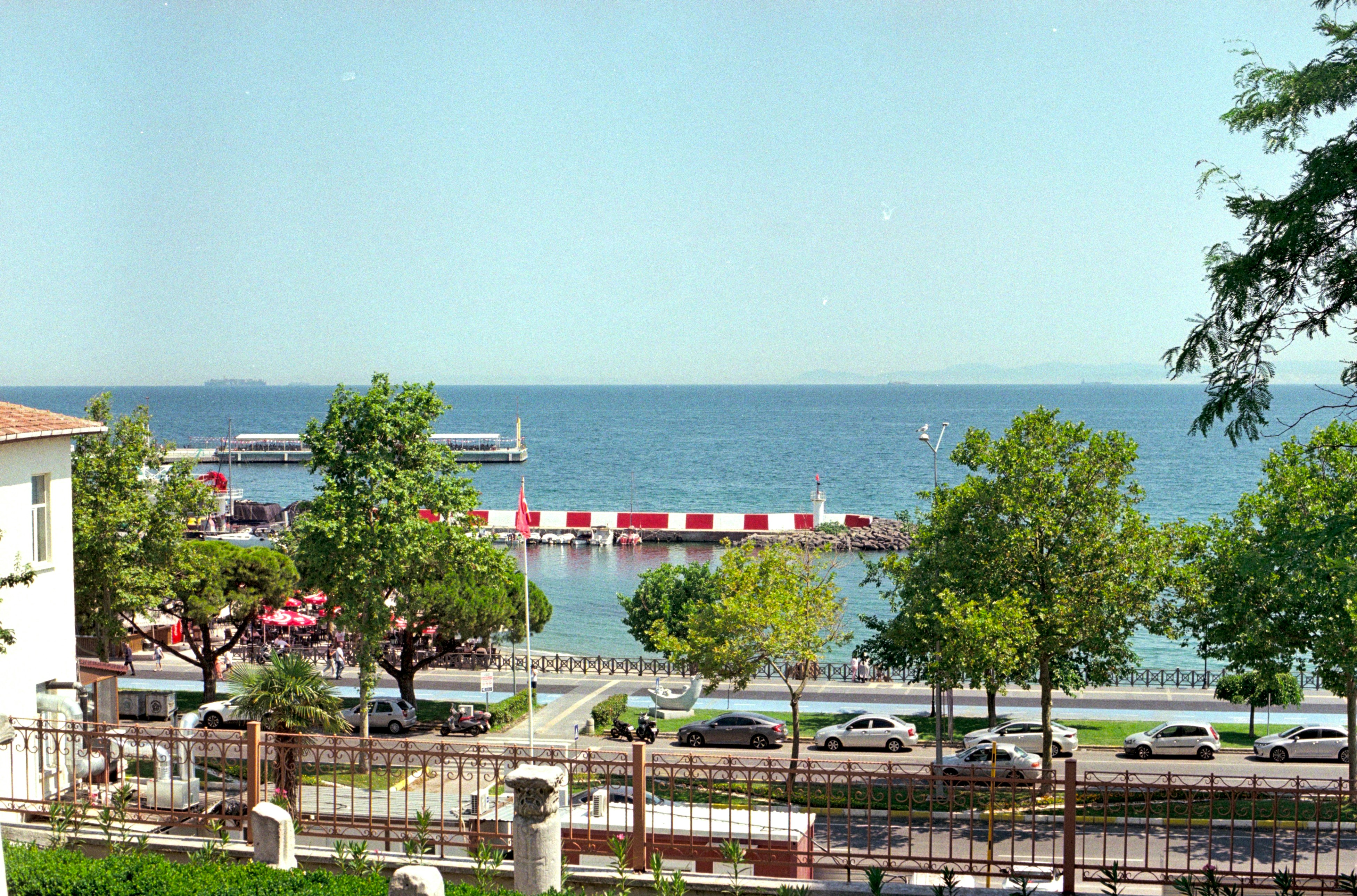 Coastal scene featuring a tree-lined promenade along a calm sea, with a ferry and distant horizon.