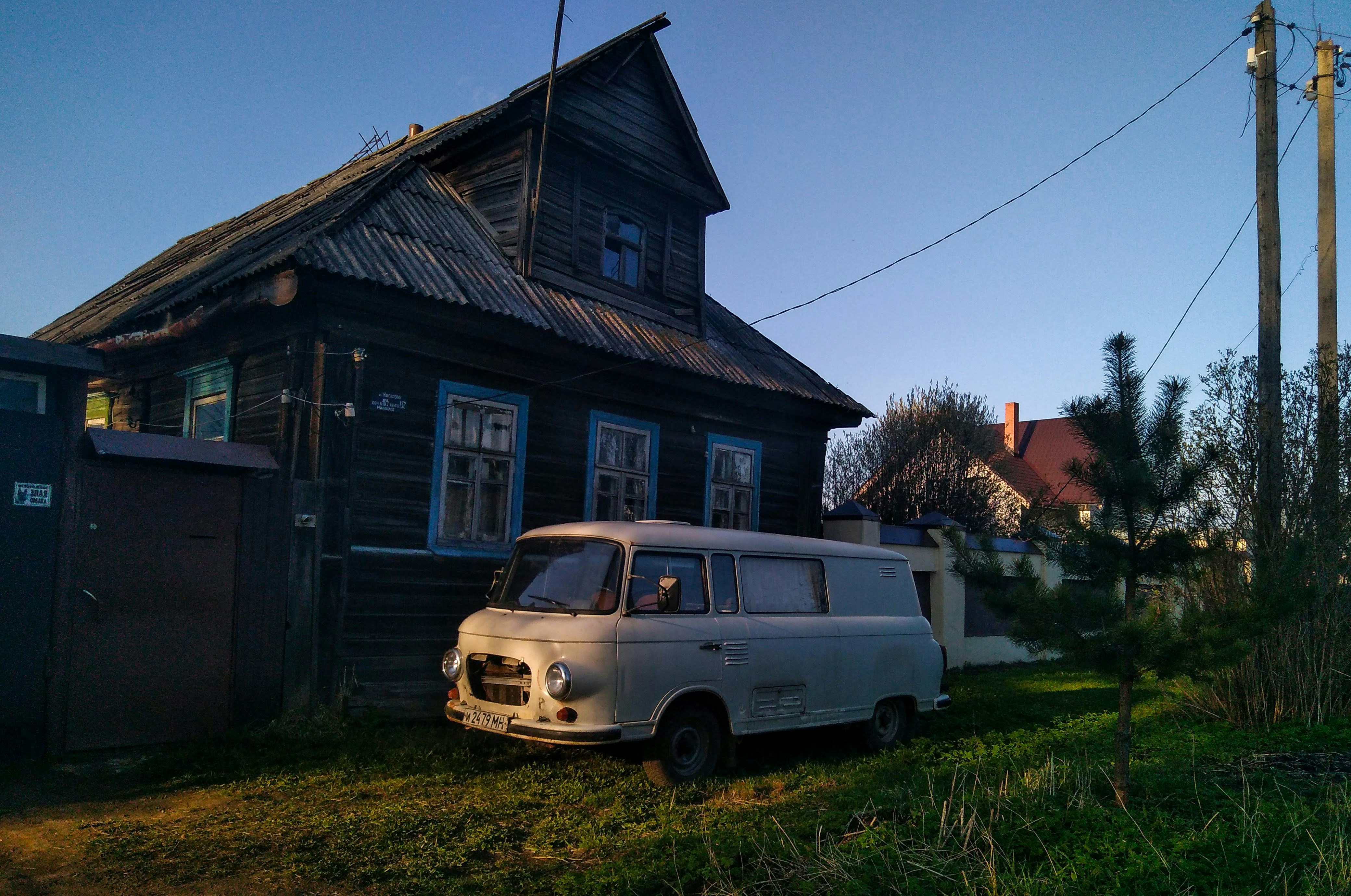 A weathered white van sits beside a dark wooden house in a quiet rural setting at dawn. The scene emphasizes rustic charm and early light on a nostalgic street.