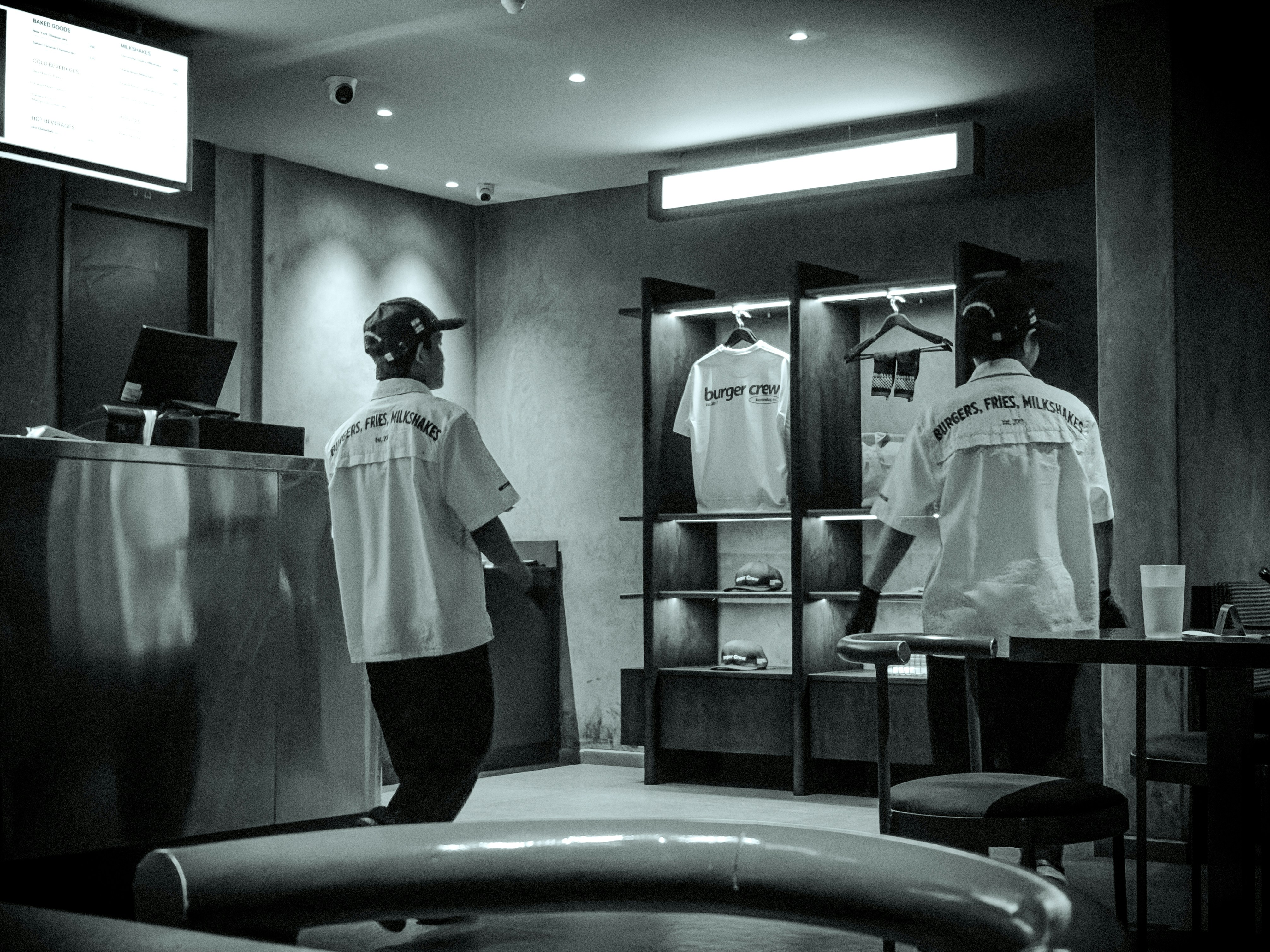 Two workers stand in a monochrome locker room, facing away from the camera near open shelves and a counter. This is a candid interior scene emphasizing uniforms and a quiet, behind-the-scenes moment.