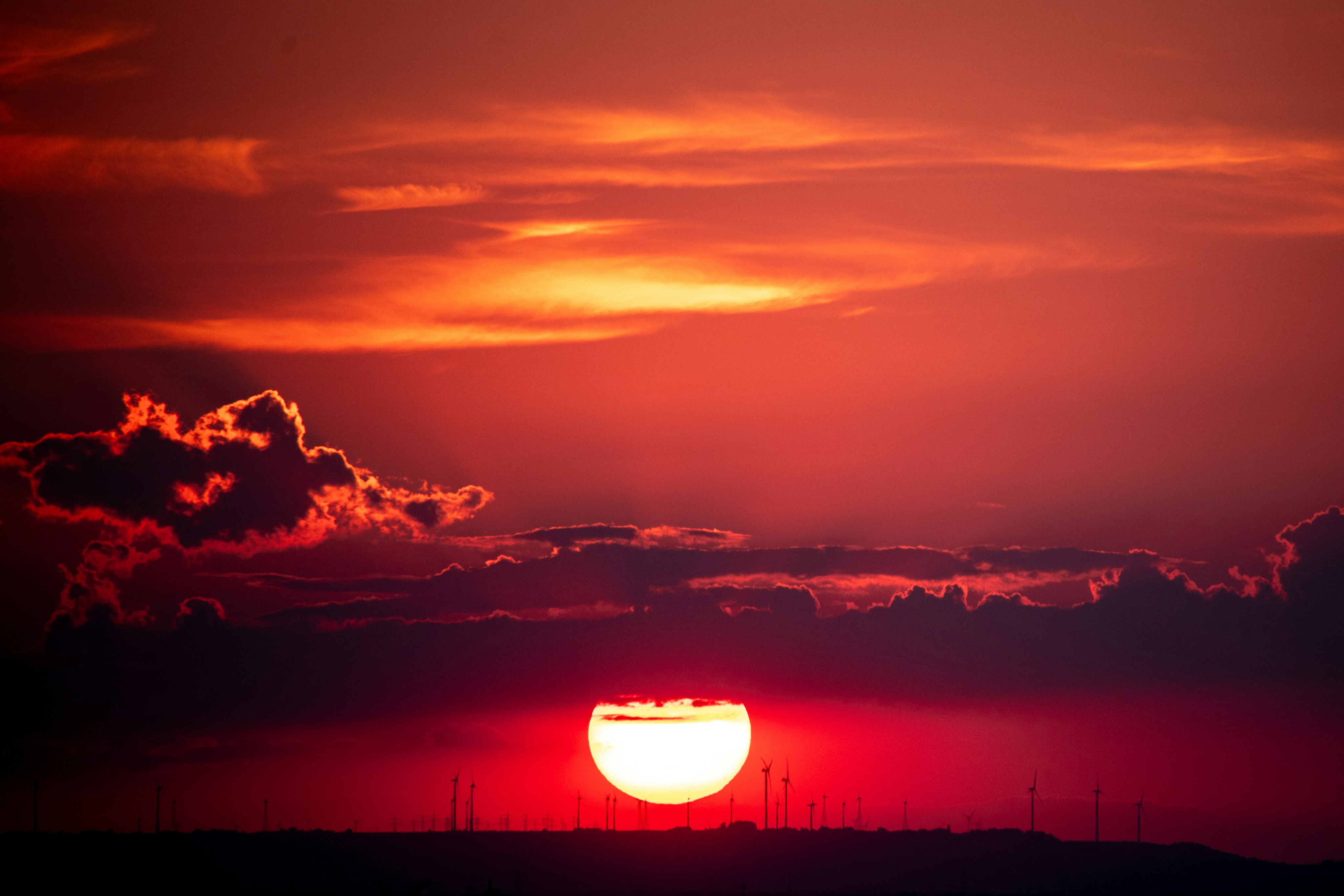 Crimson Horizon: The Sun Dips Behind a Silhouette of Distant Wind Turbines
