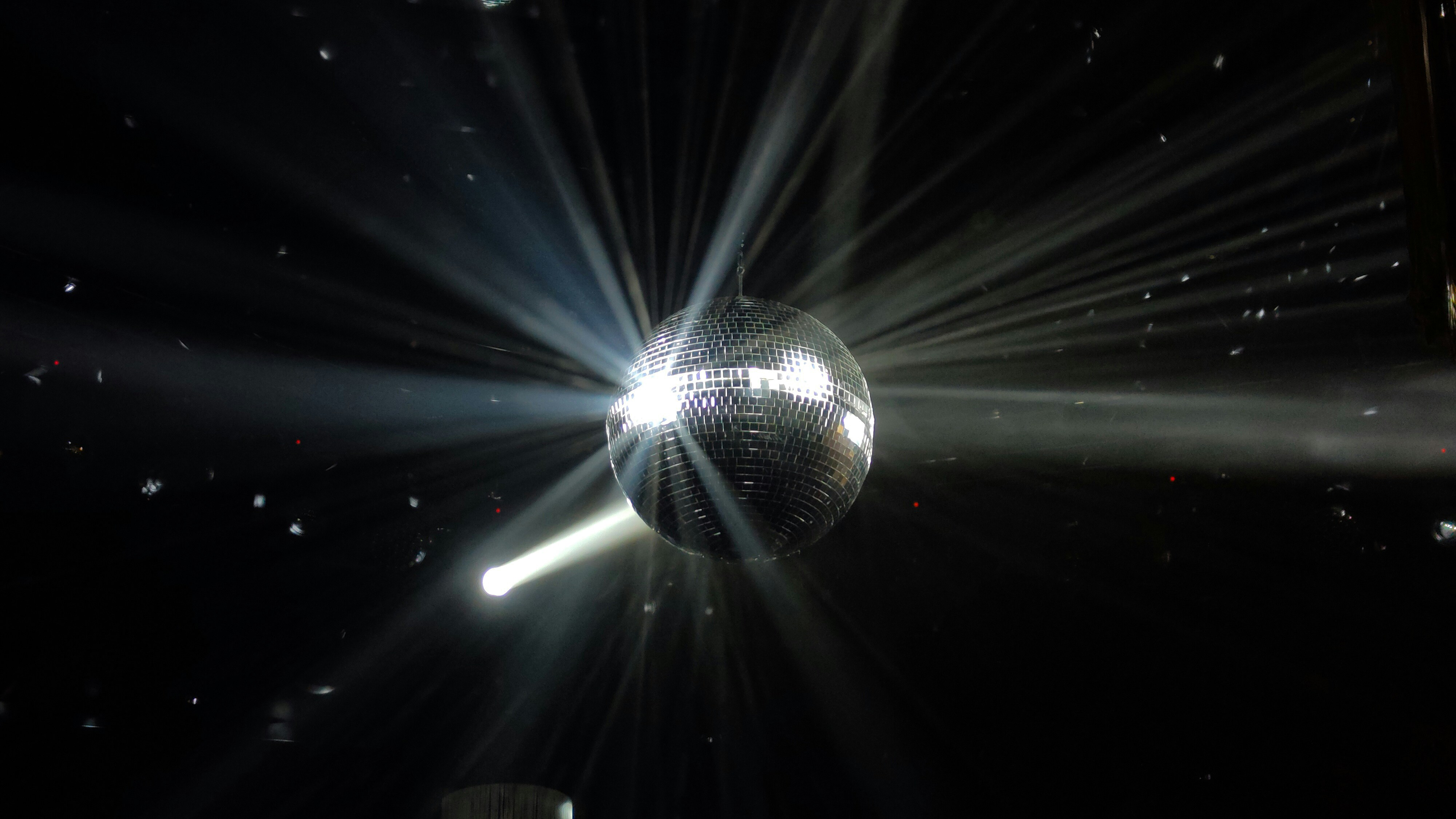 A mirrored disco ball radiates light beams in a dark space, surrounded by specks of confetti-like particles and theatrical beams. The scene centers on the orb, suggesting a performance or party atmosphere.