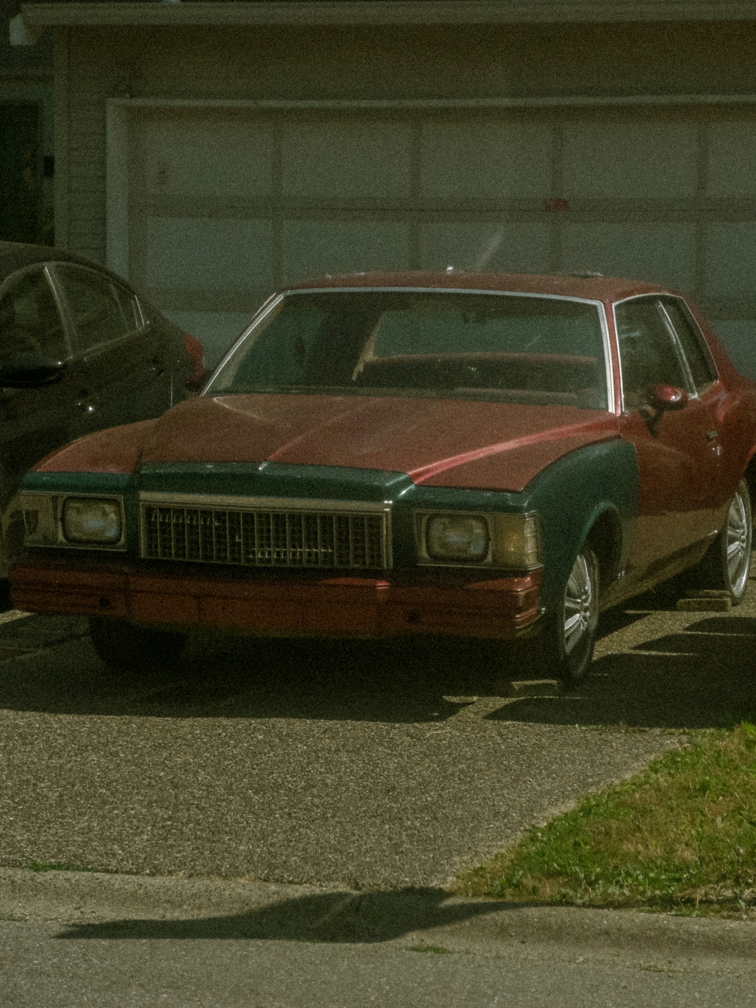 Red and green vintage car parked in a driveway beside a closed garage, with muted daylight and a neighboring black vehicle. A small patch of grass and pavement slabs anchor the scene.