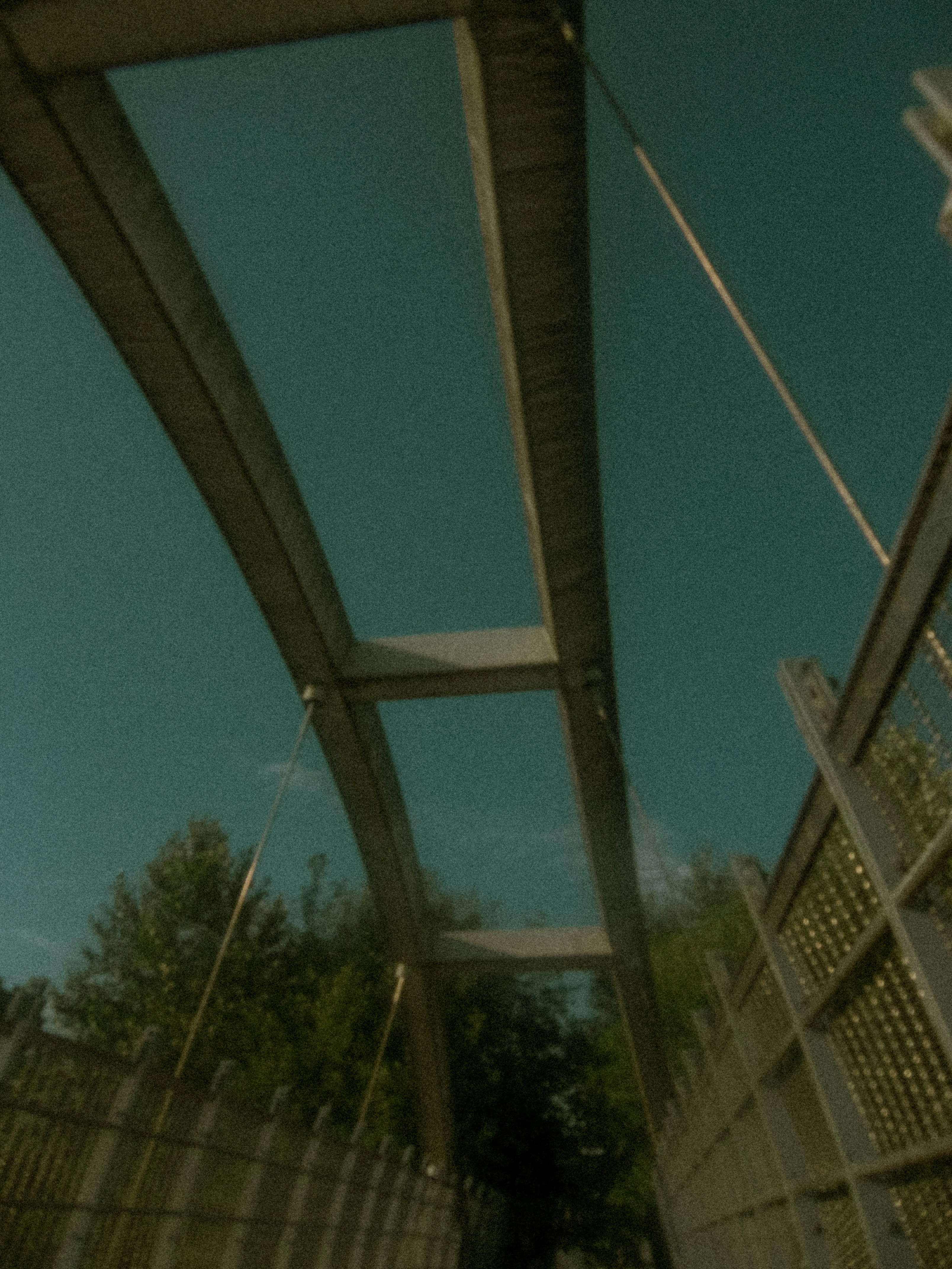 Industrial walkway over a green-turred sky framed by steel beams and wire rails, suggesting a pedestrian bridge or skyway. This is a candid architectural scene.