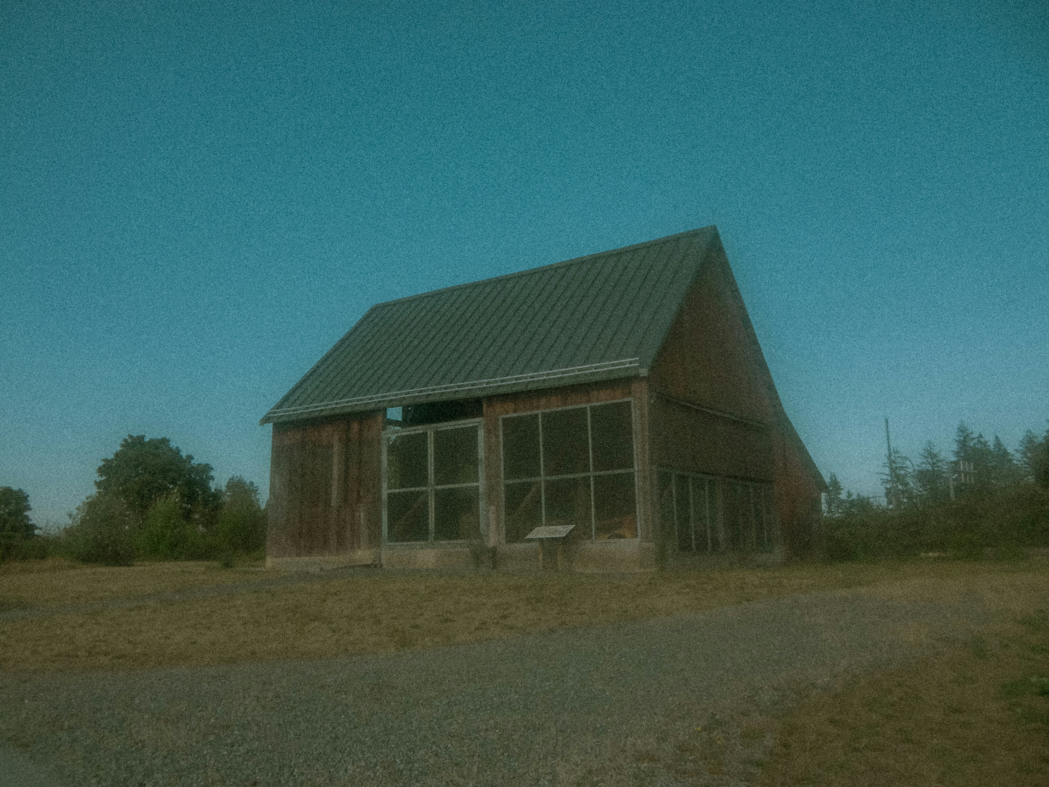 Weathered barn amid open field under blue sky with a dirt road foreground, softly lit and slightly hazy.
