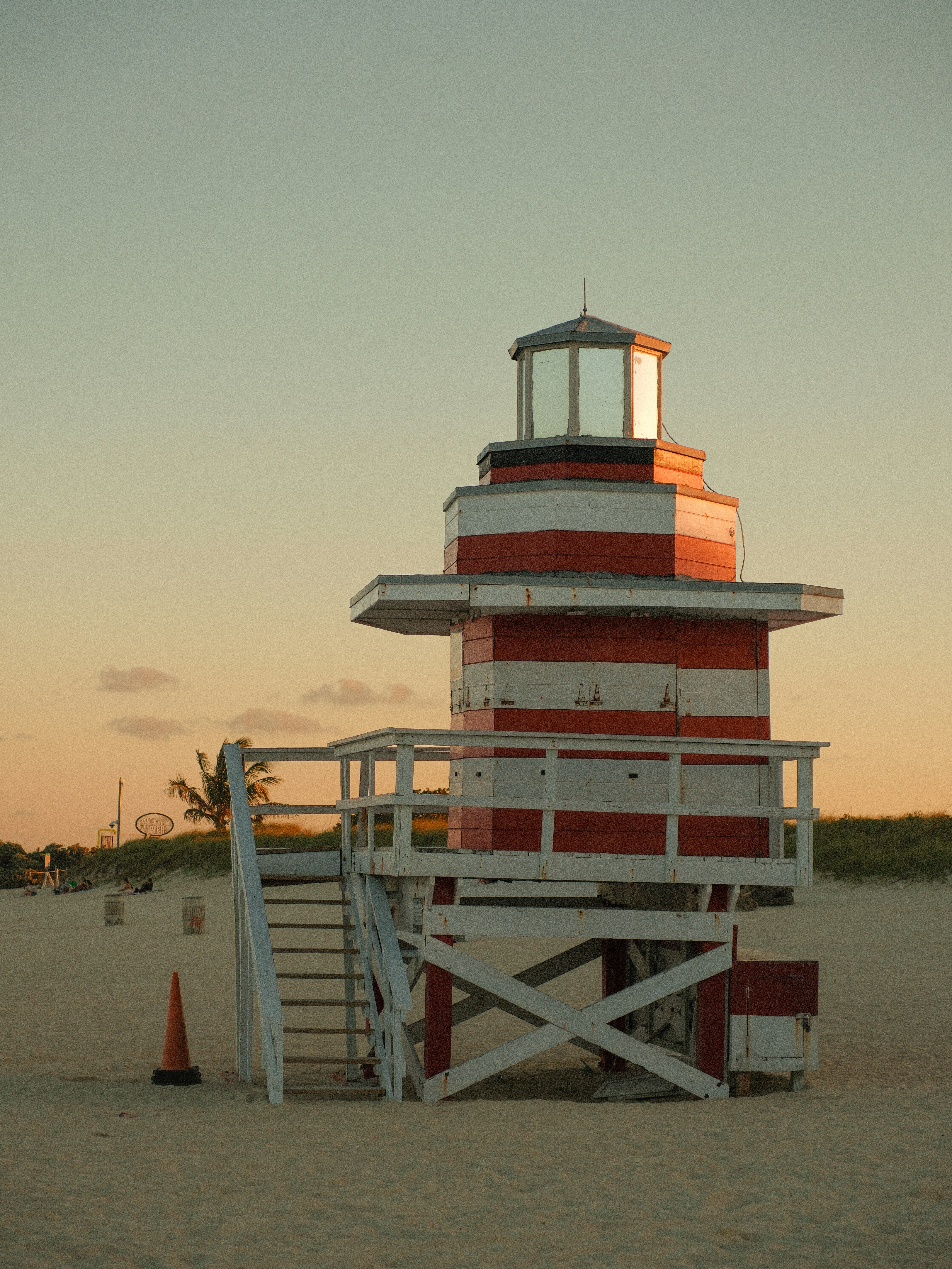 A red and white lifeguard tower sits on a sandy beach during golden hour, with soft light shaping the weathered wood and calm sky in the background.