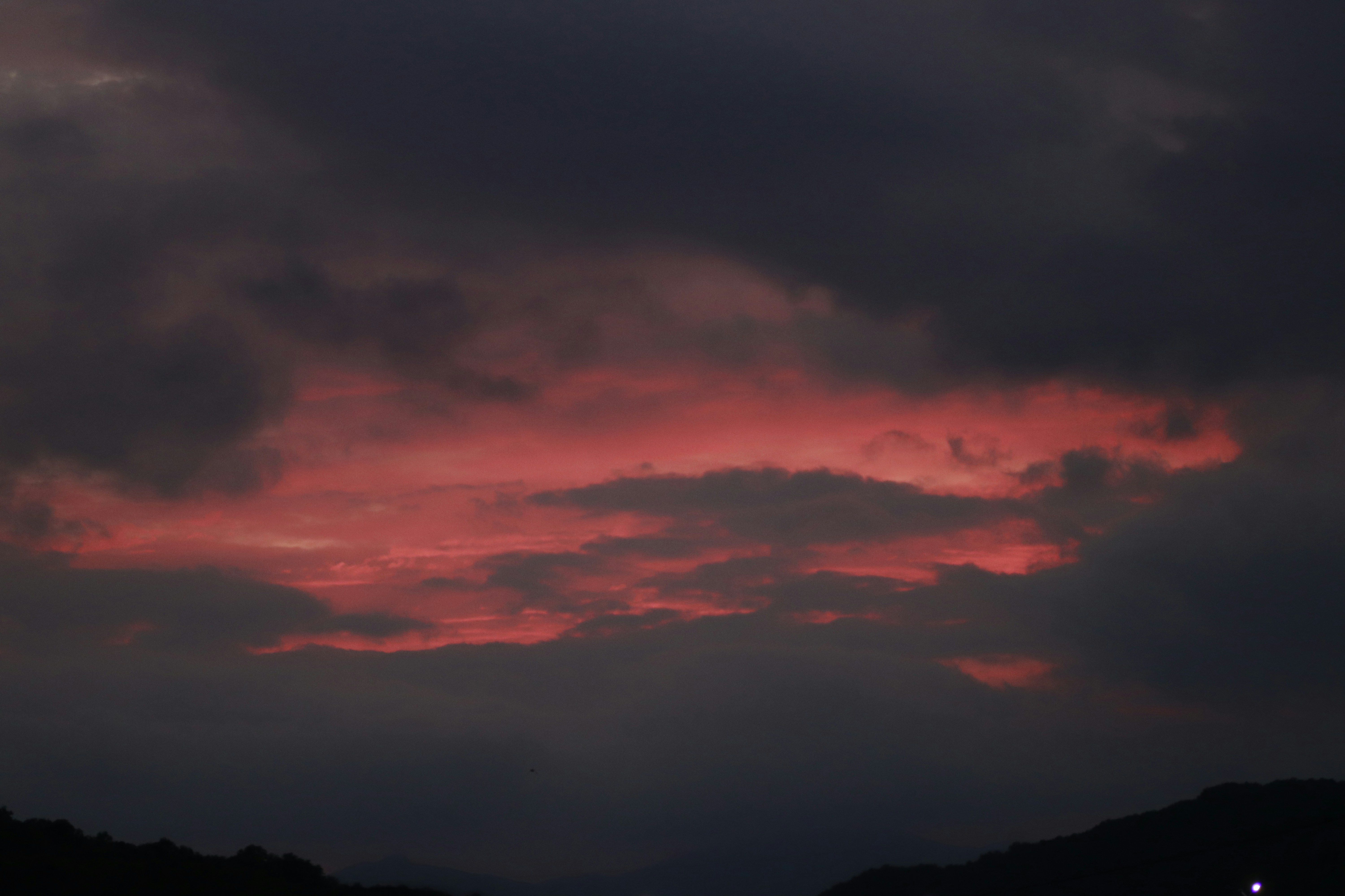 Burnt-red clouds glow above a dark horizon as twilight settles, silhouette of distant hills under a moody sky. This is a landscape shot emphasizing atmospheric color and quiet drama.