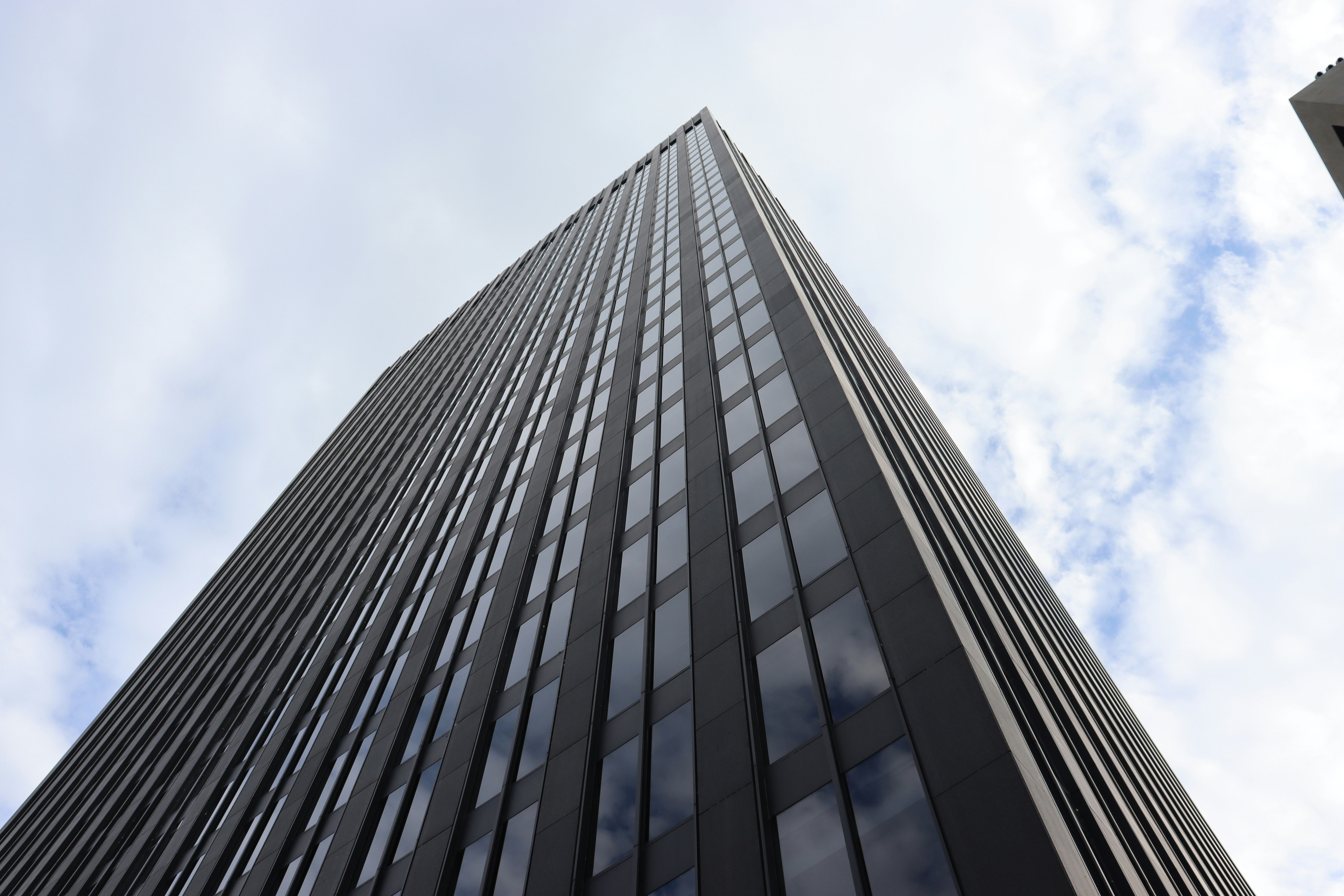 Tall glass-and-steel skyscraper tilts upward with clouds in the background, emphasizing architectural lines and vertical scale. A stark, modern structure captured from a low-angle perspective.