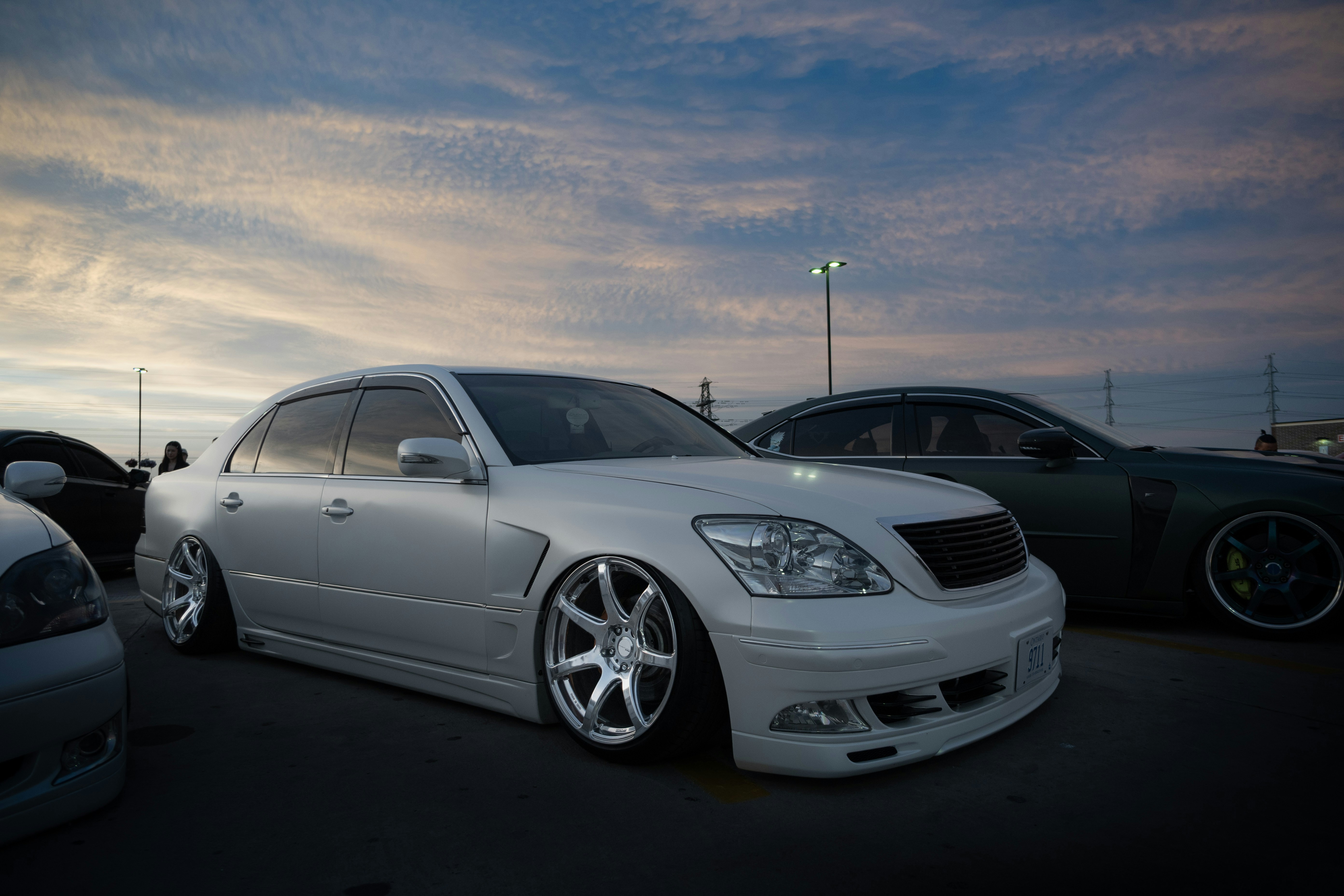 Low-angle shot of a white modified sedan with large chrome wheels in a parking lot, sporty car ahead of other vehicles; subdued sunset lighting highlights the glossy paint.