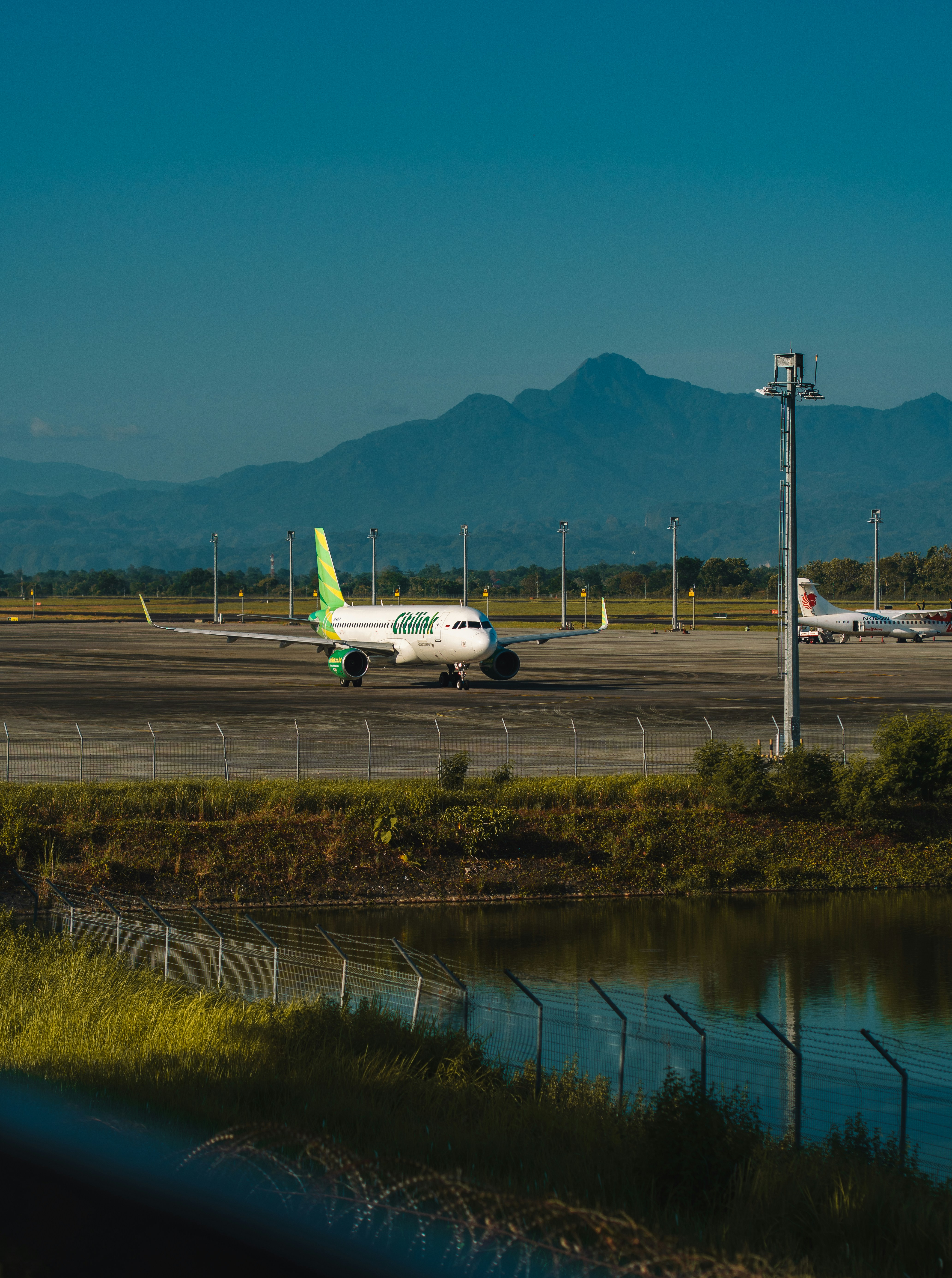 A green-tailed airplane sits on a taxiway at a quiet airport, with hangars and utility poles beyond and a distant mountain range under a clear sky.