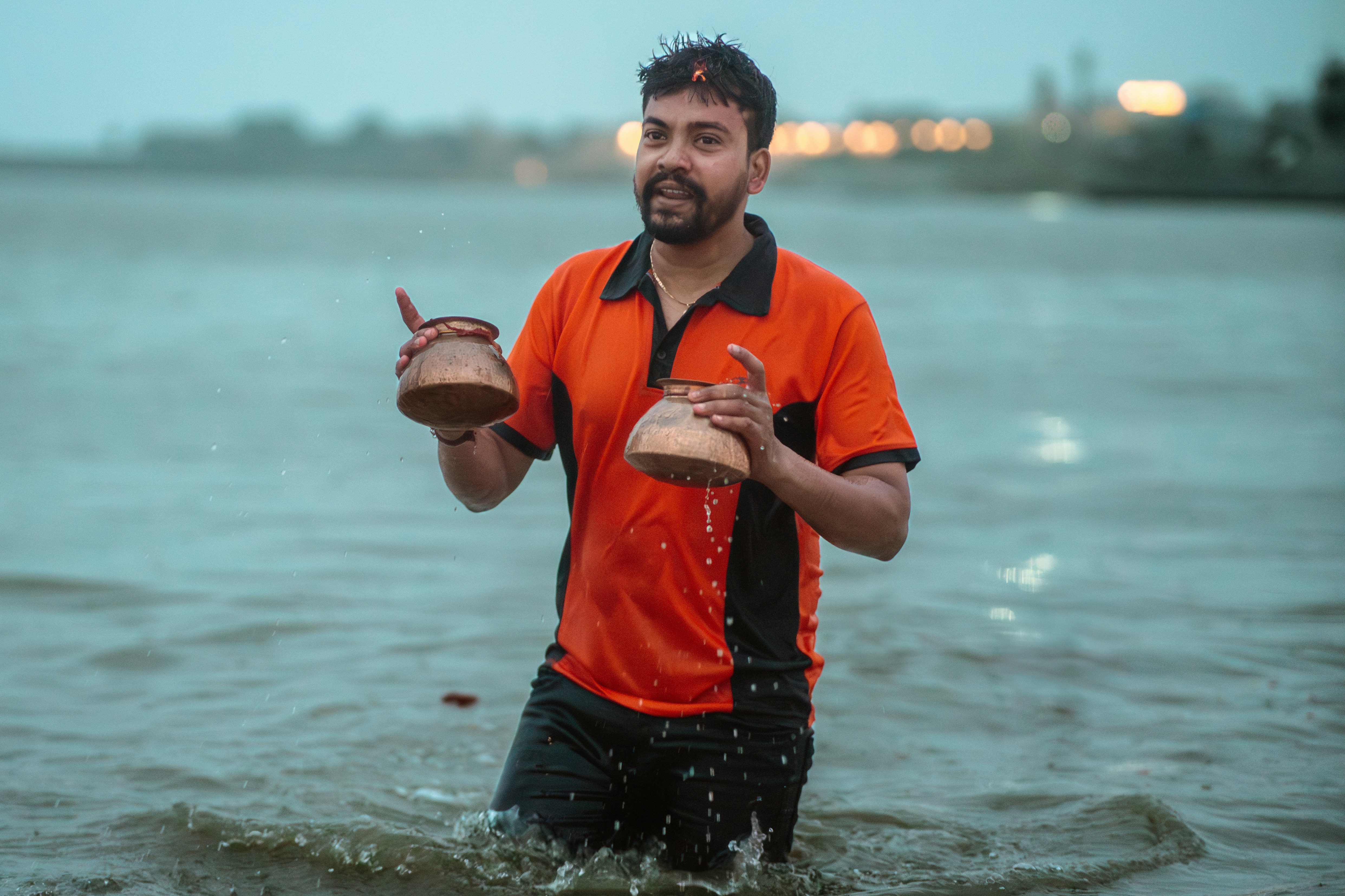 Aman in orange at the shoreline, cups in hand, bathed in ocean spray