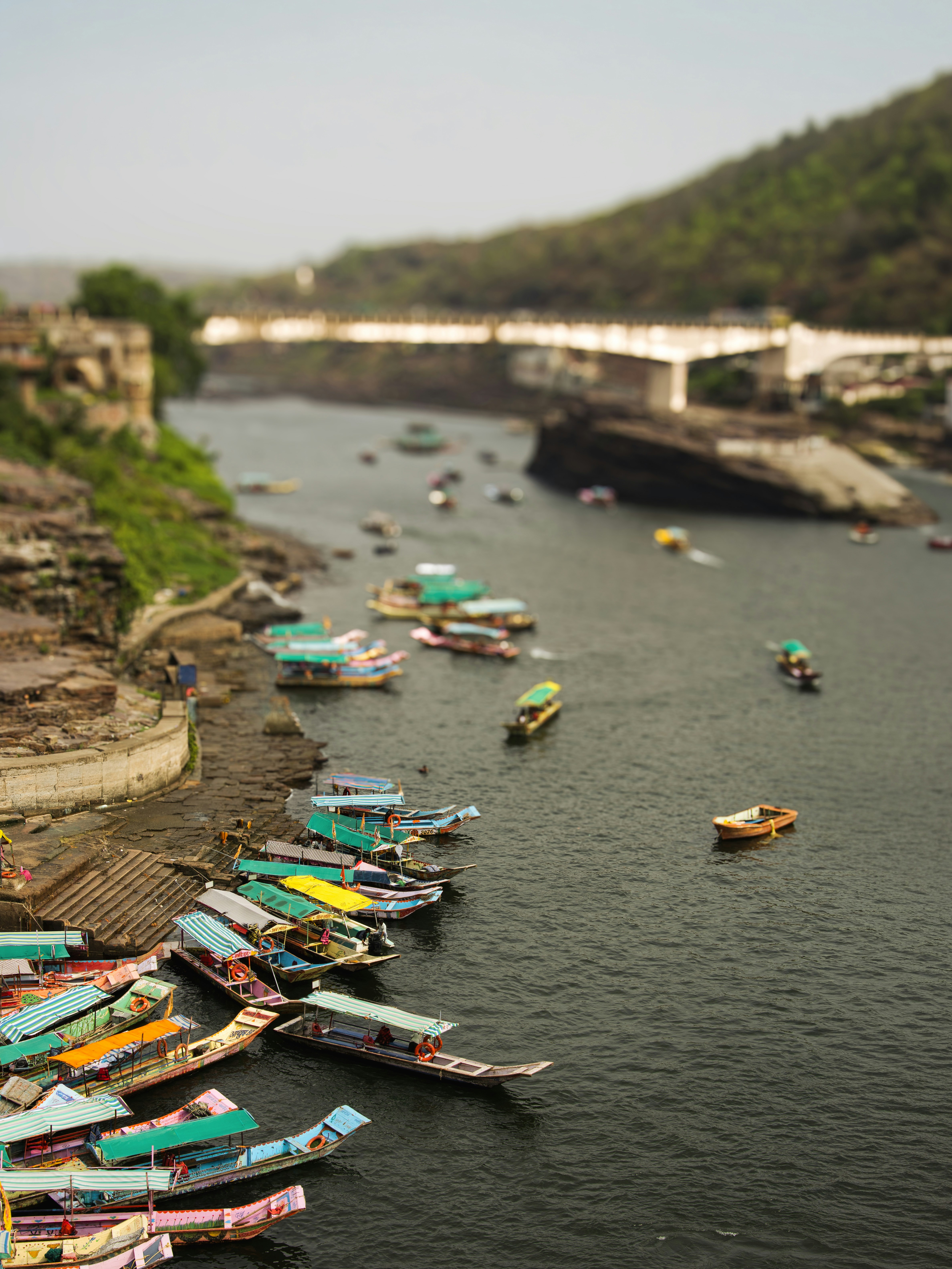 Scenic view of the Narmada River in the holy Hindu temple town of Omkareshwar, Madhya Pradesh, India. The photo shows colorful wooden tourist boats docked at the stone steps (ghats) with a suspension bridge and hills in the background, captured with a creative tilt-shift blur.