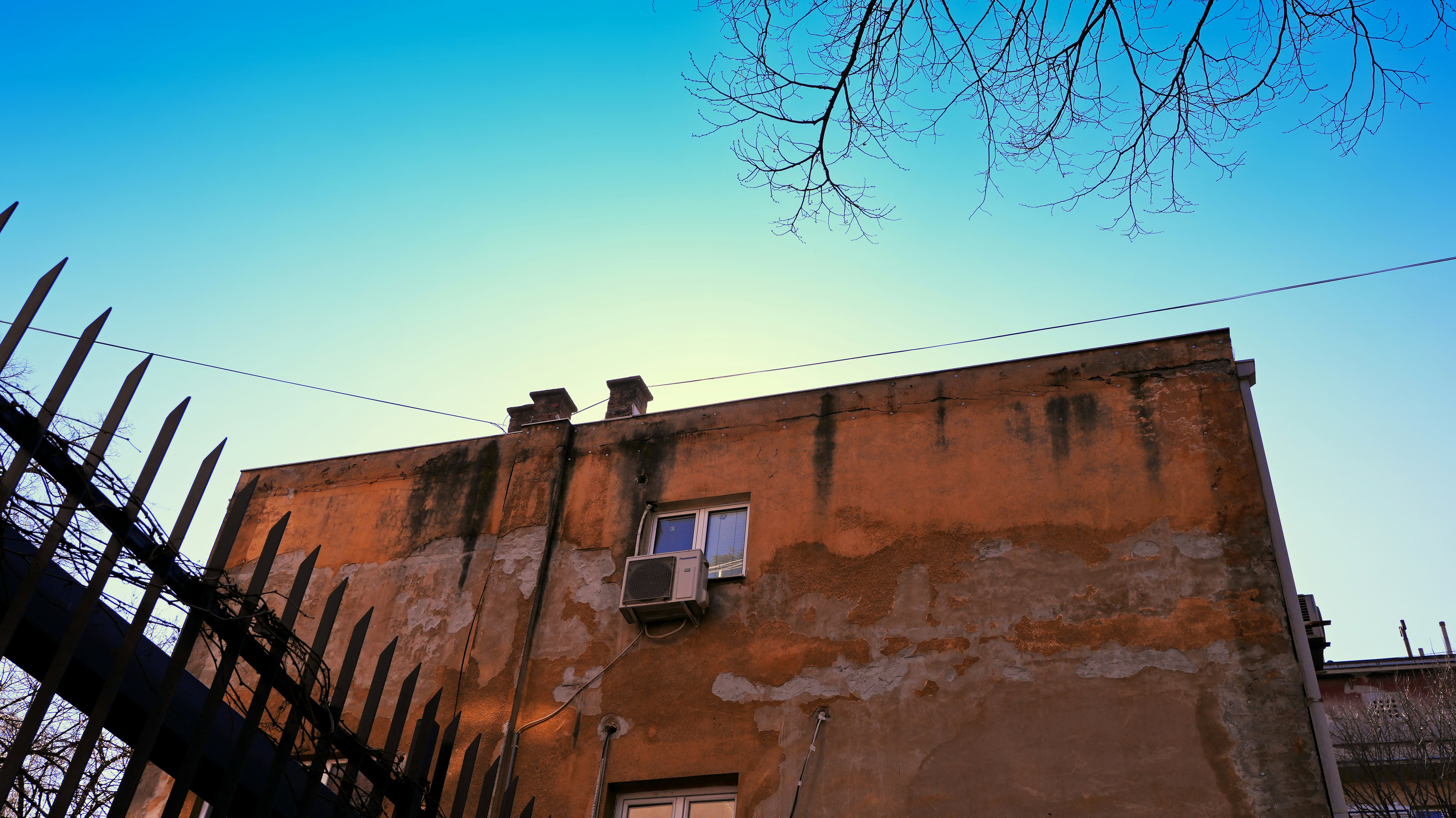Brick building wall with peeling plaster and visible air conditioner unit, under a clear blue sky; a portion of a fence is visible in the left foreground. A simple architectural detail shot emphasizing texture and color.