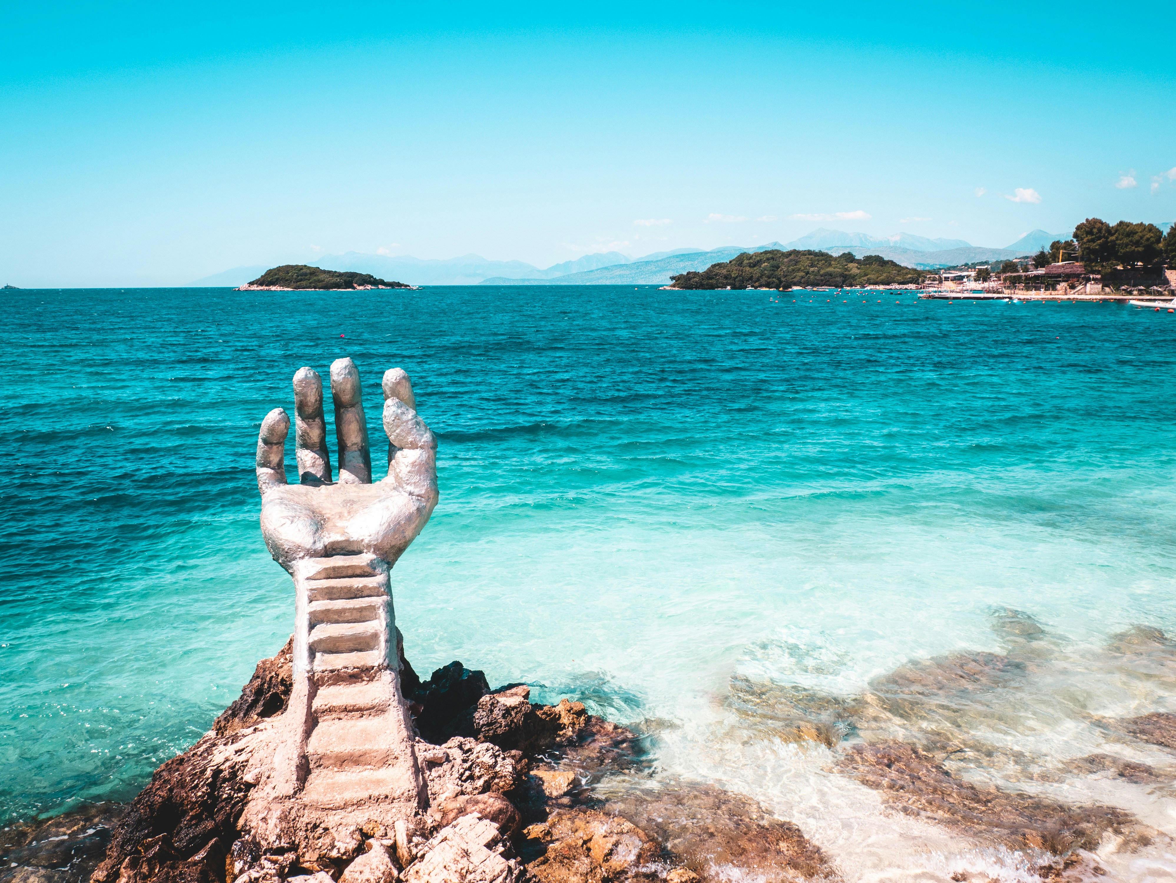 A stone hand statue rising from rocky shore reaches toward turquoise water, with a calm sea and distant islands on a bright day. a coastal scene with a sculpted hand as the focal point.