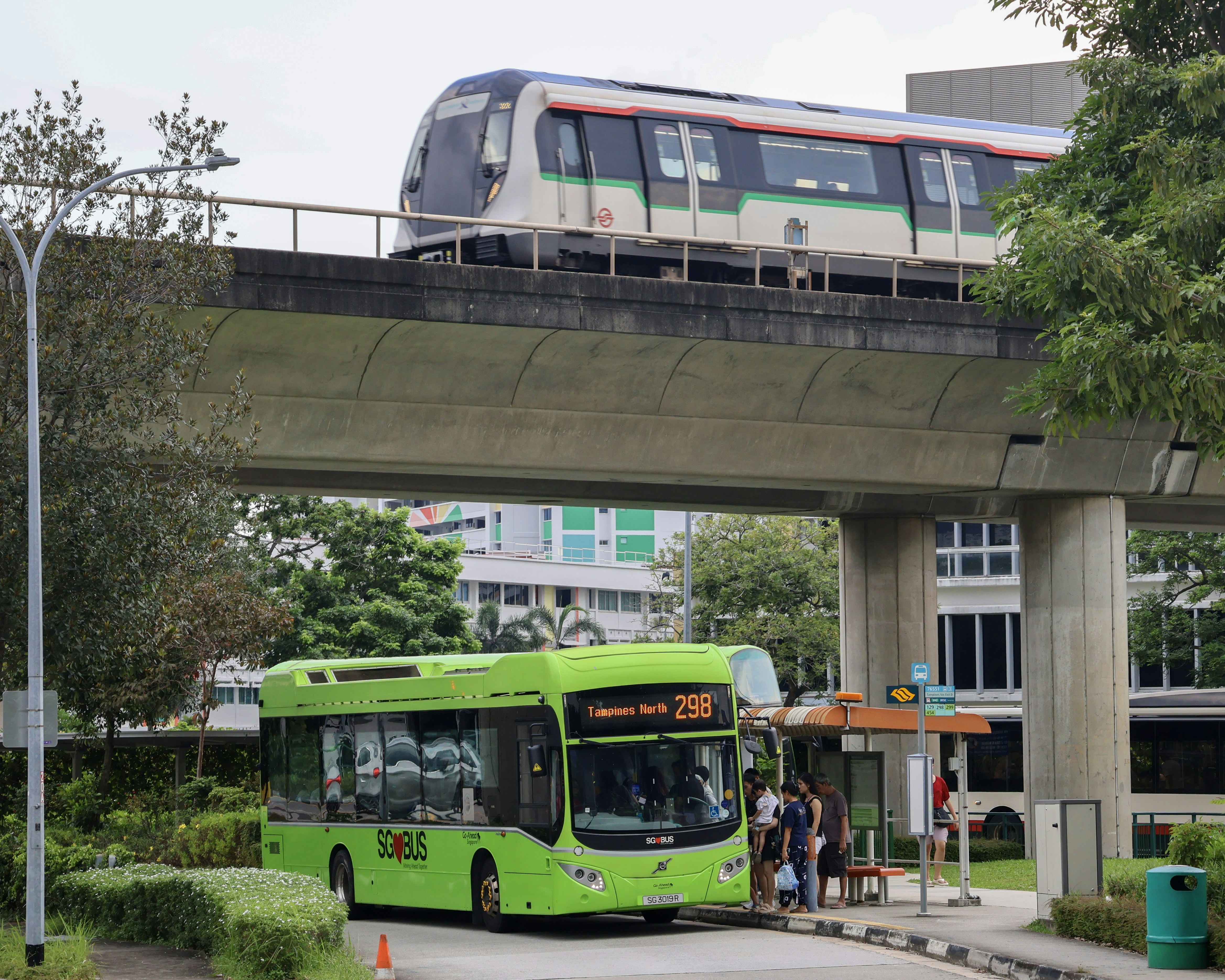 Green city bus at a stop beneath an elevated rail line, with passengers waiting nearby and a modern train passing overhead.