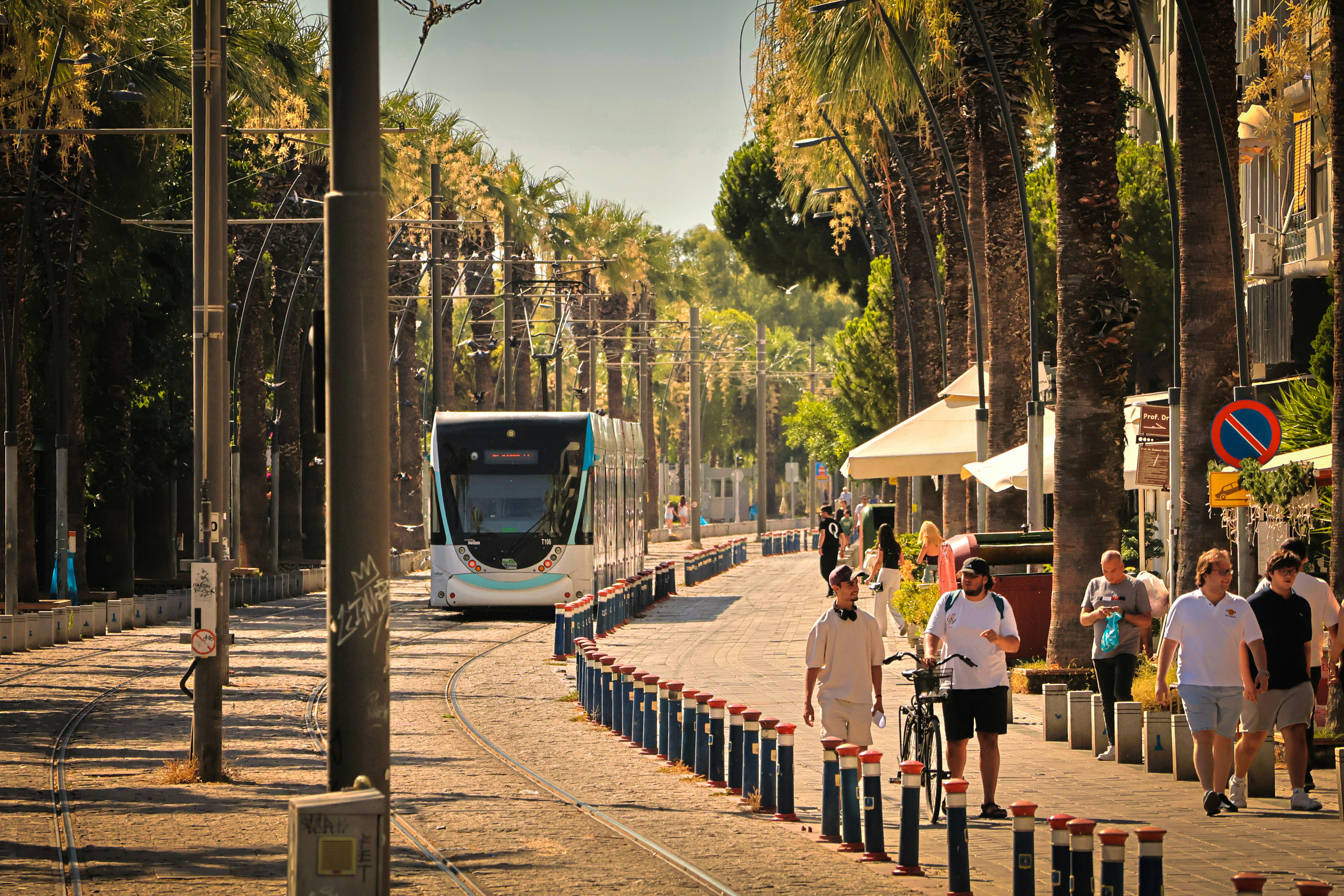 A sunlit city street with a modern tram approaching, lined by palm trees and pedestrians along a pedestrian-friendly boulevard.