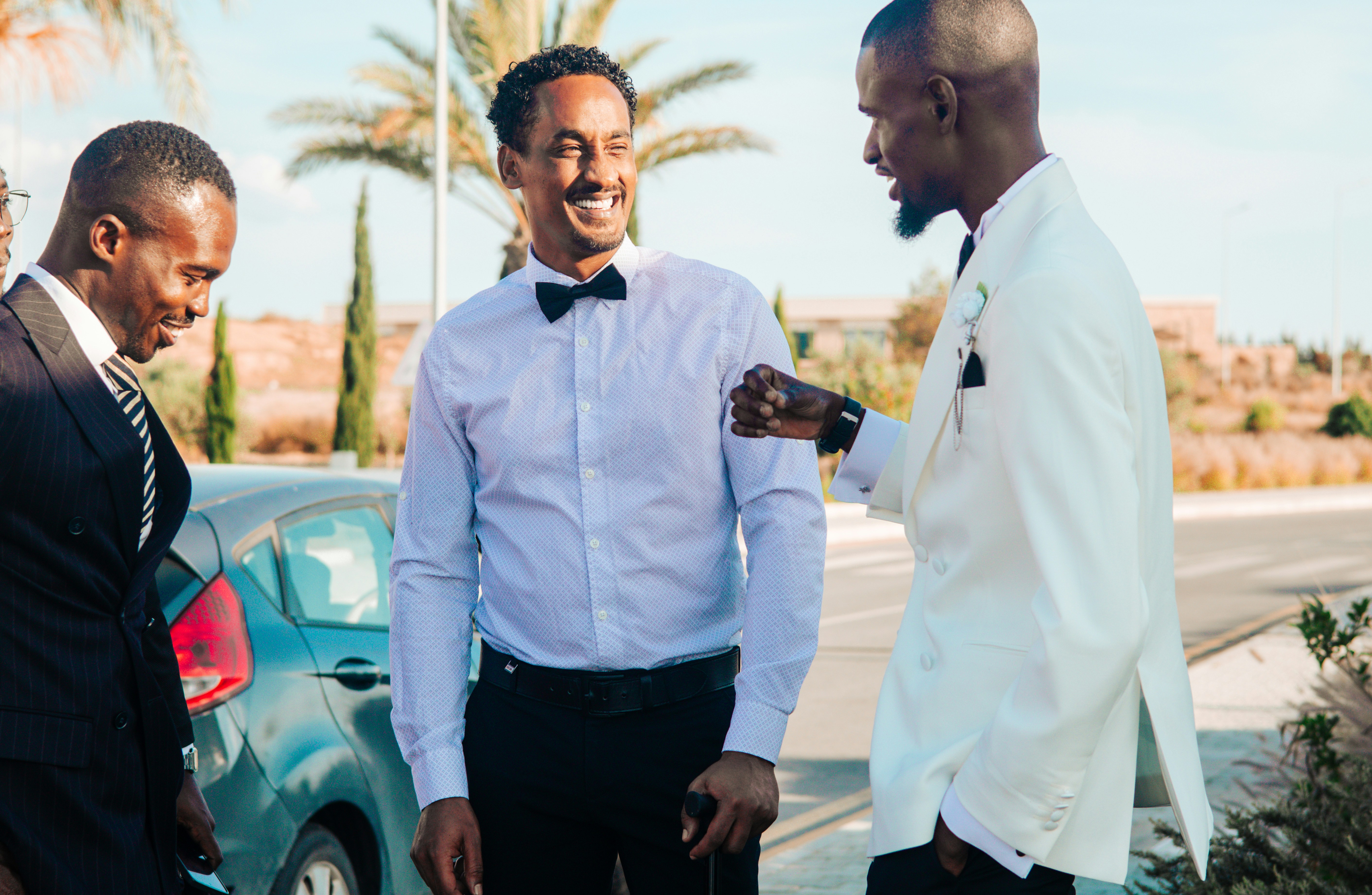 Groomed men share a lighthearted moment outside, dressed in formal attire with a car and sunny backdrop in the scene.