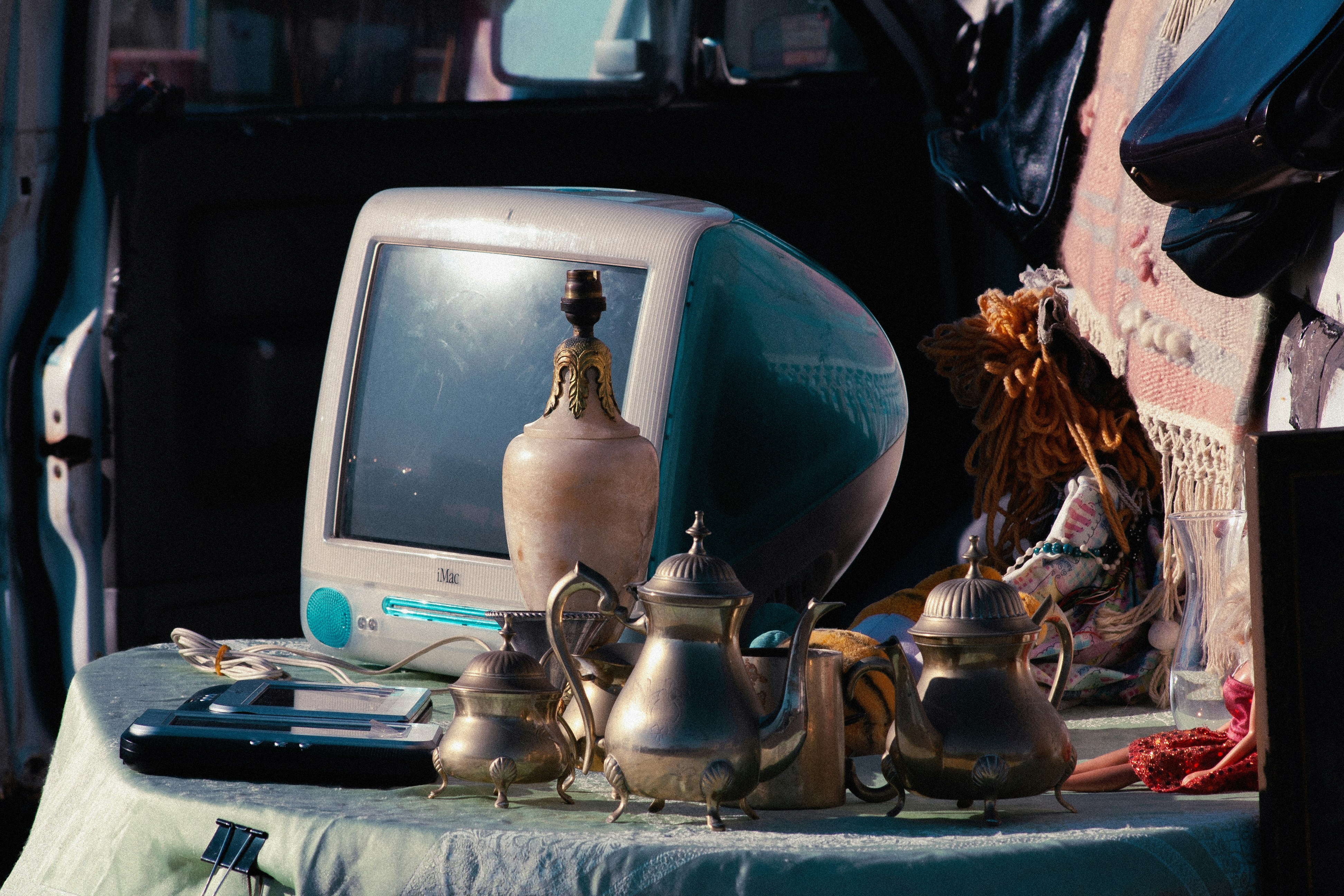 A vintage still-life scene with an old CRT monitor and a row of tarnished teapots on a teal-covered table, accompanied by decorative textiles in the background.