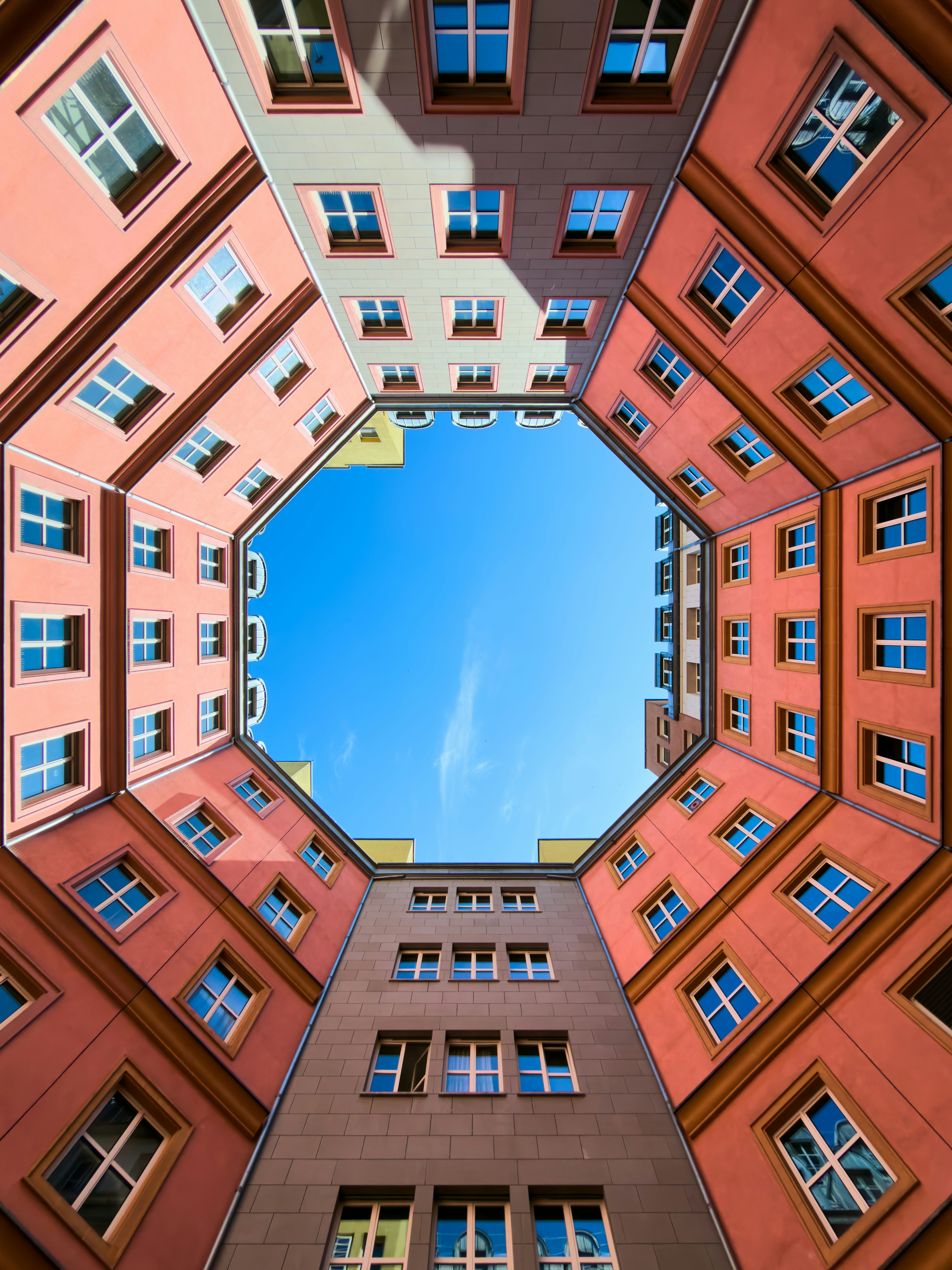 A square-formed courtyard of salmon and peach-hued buildings opens to a bright blue sky, captured from the center of the atrium with windows forming a geometric frame.