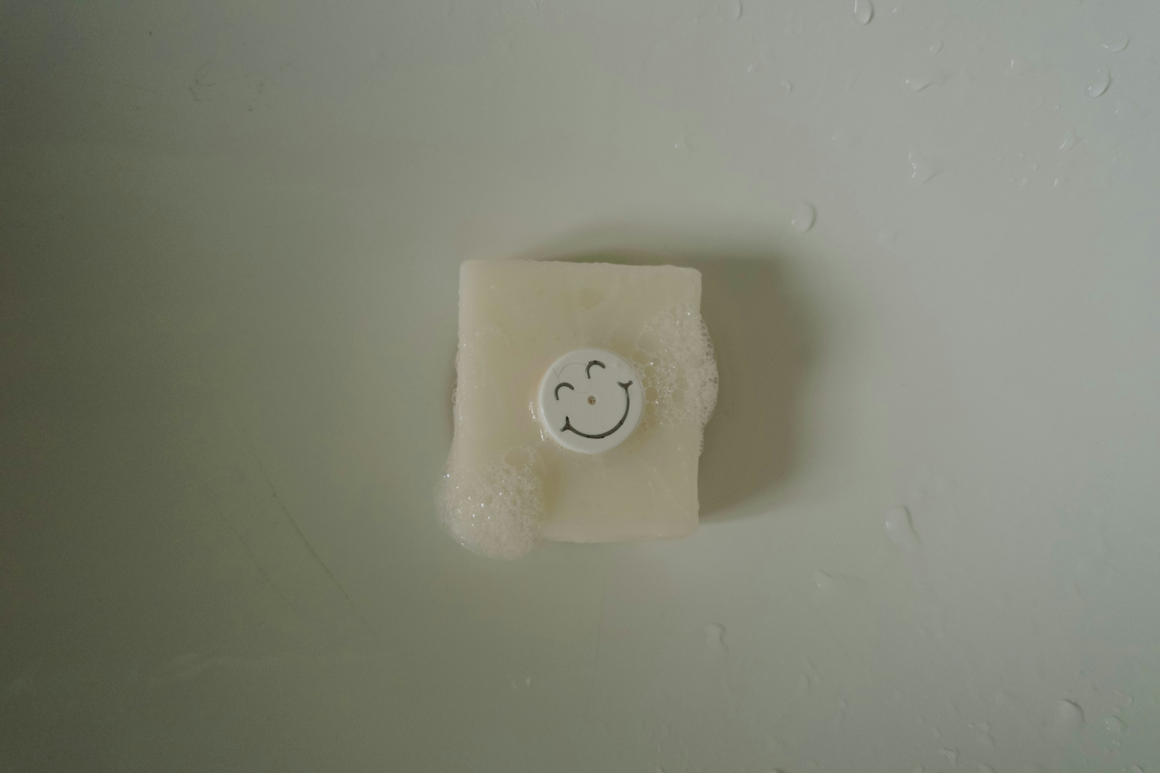 A small piece of soap with a hand-drawn smiley square rests on a pale, wet surface; the tiny subject conveys a playful focal point amidst a minimalist background.