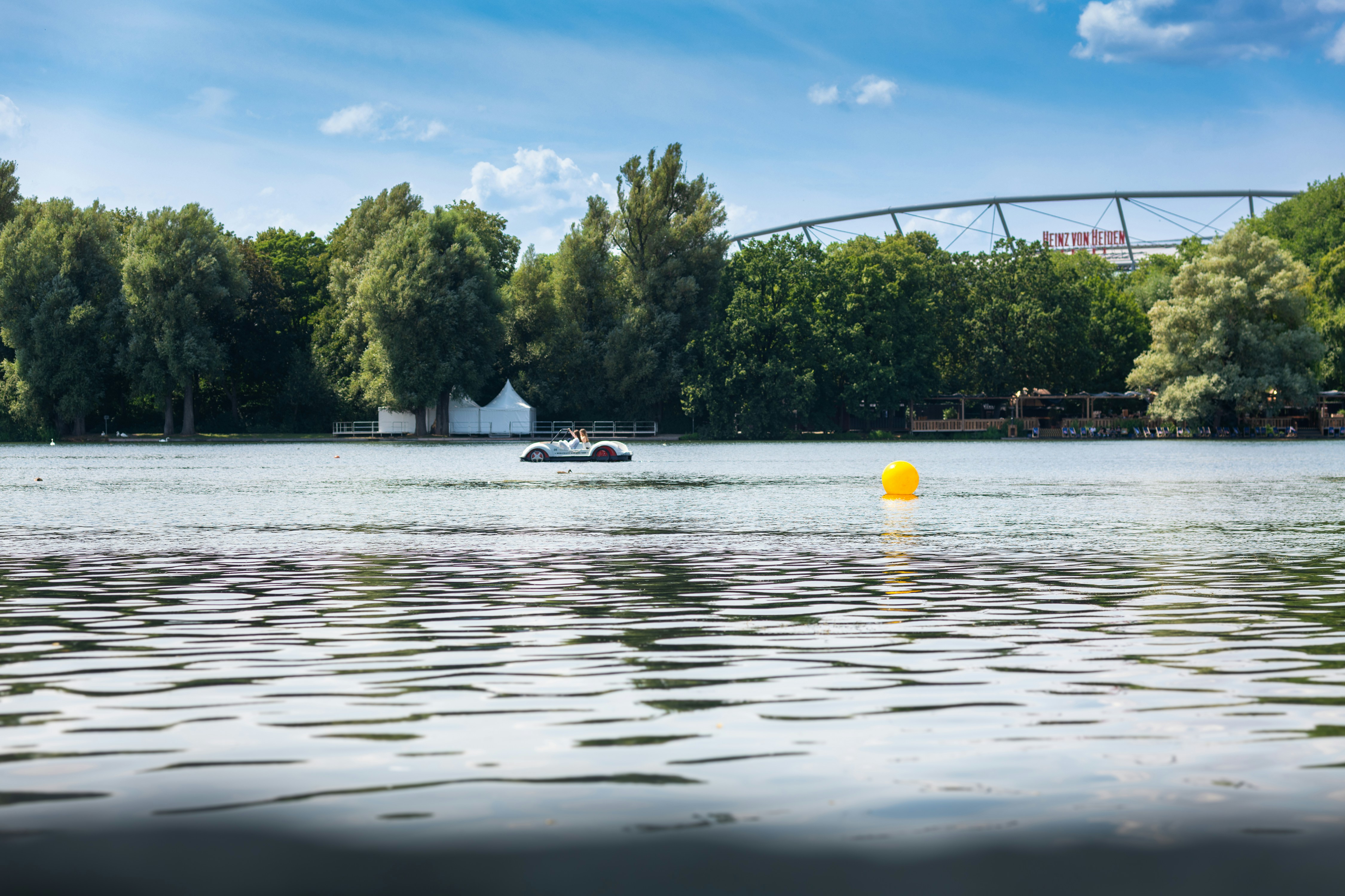 Calm lake scene with a small boat near the tree-lined shore and a bright yellow buoy, under a clear blue sky.