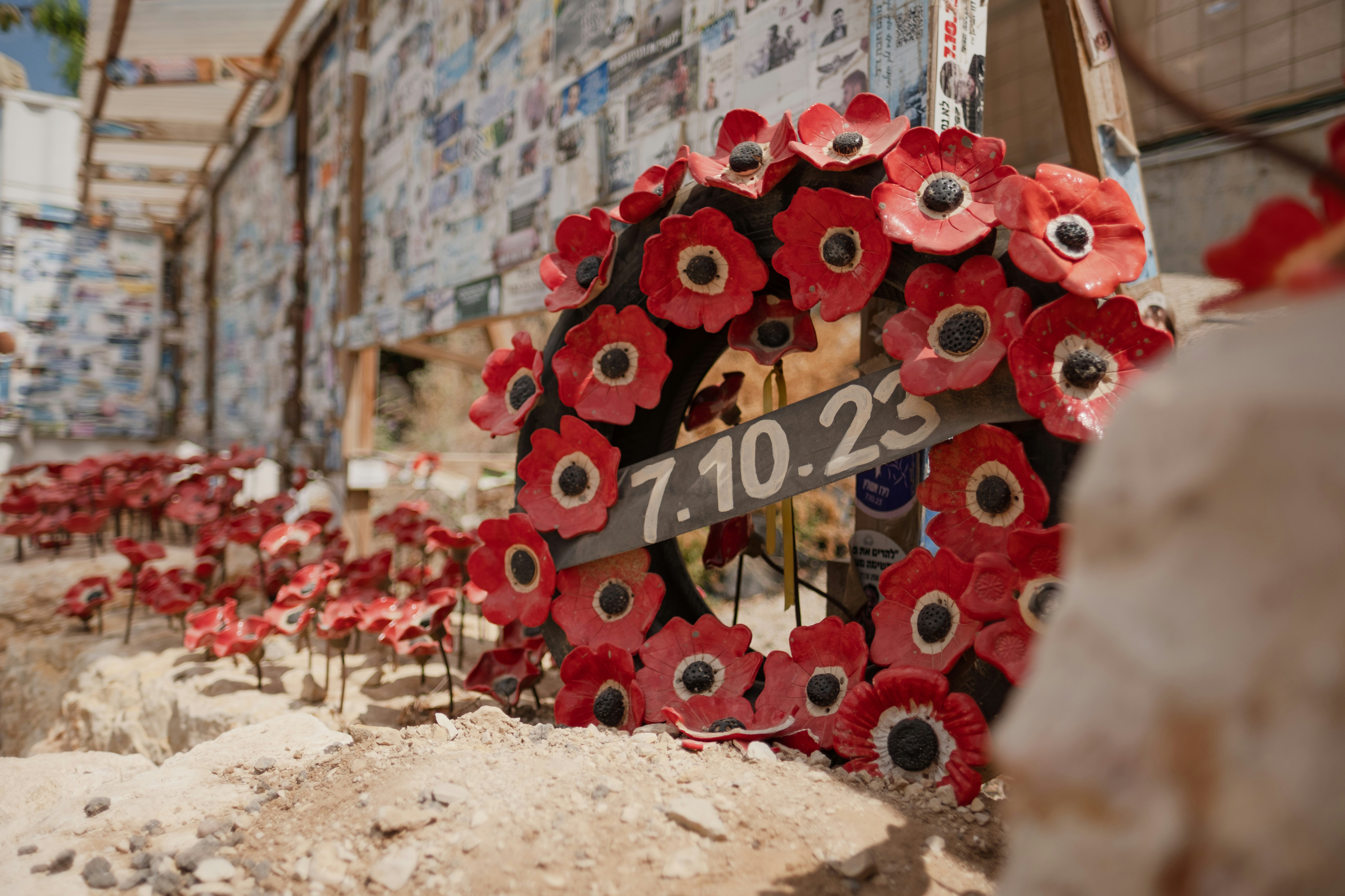 Red floral wreath with a black placard at a dusty construction site, surrounded by scattered debris and background memorial elements.
