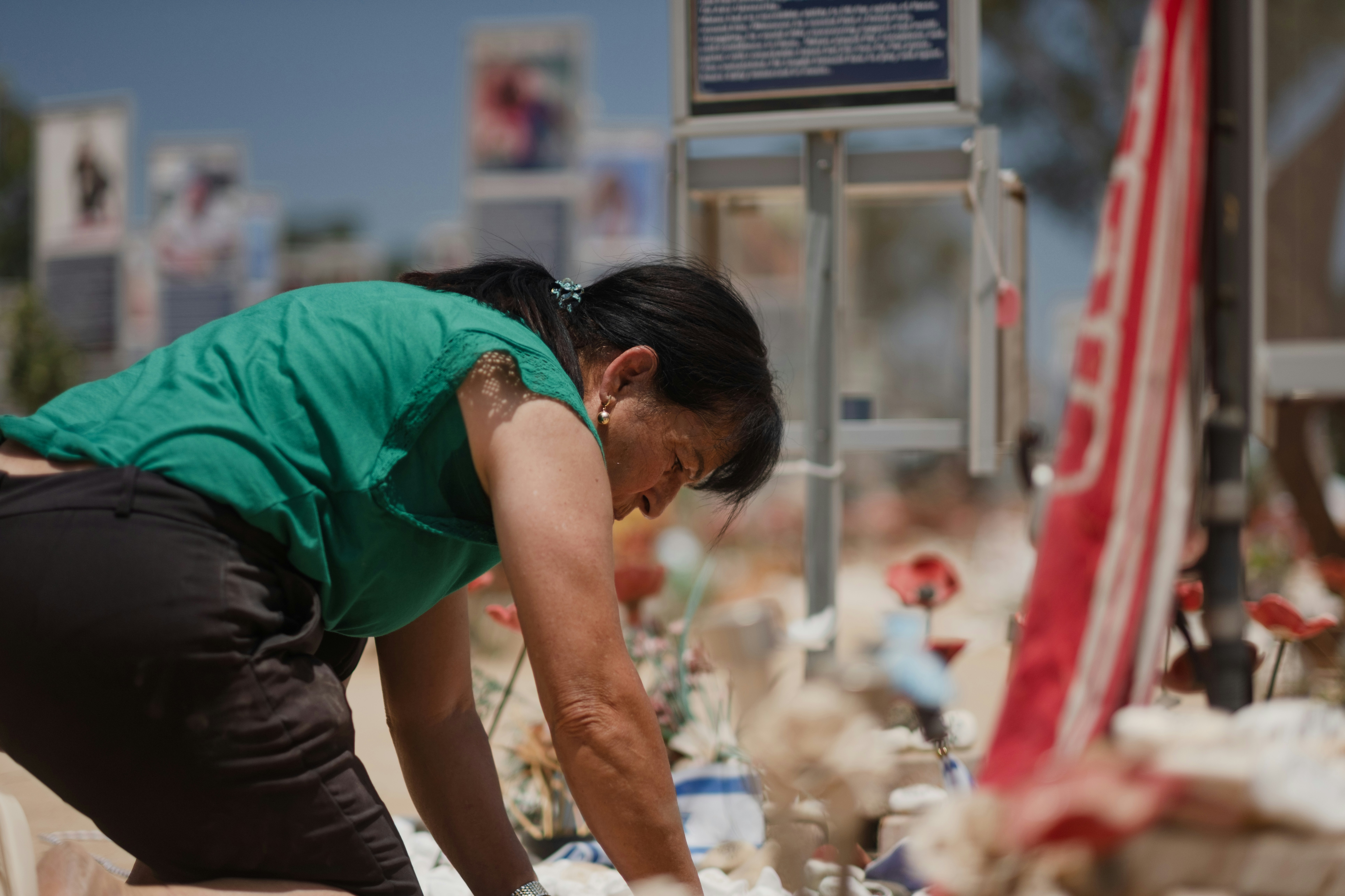A woman in a green shirt, bending over, arranges small objects at a seaside market; the scene captures a focused moment among stalls and decorative items. A logo-like flag and blurred background indicate a bustling outdoor market.