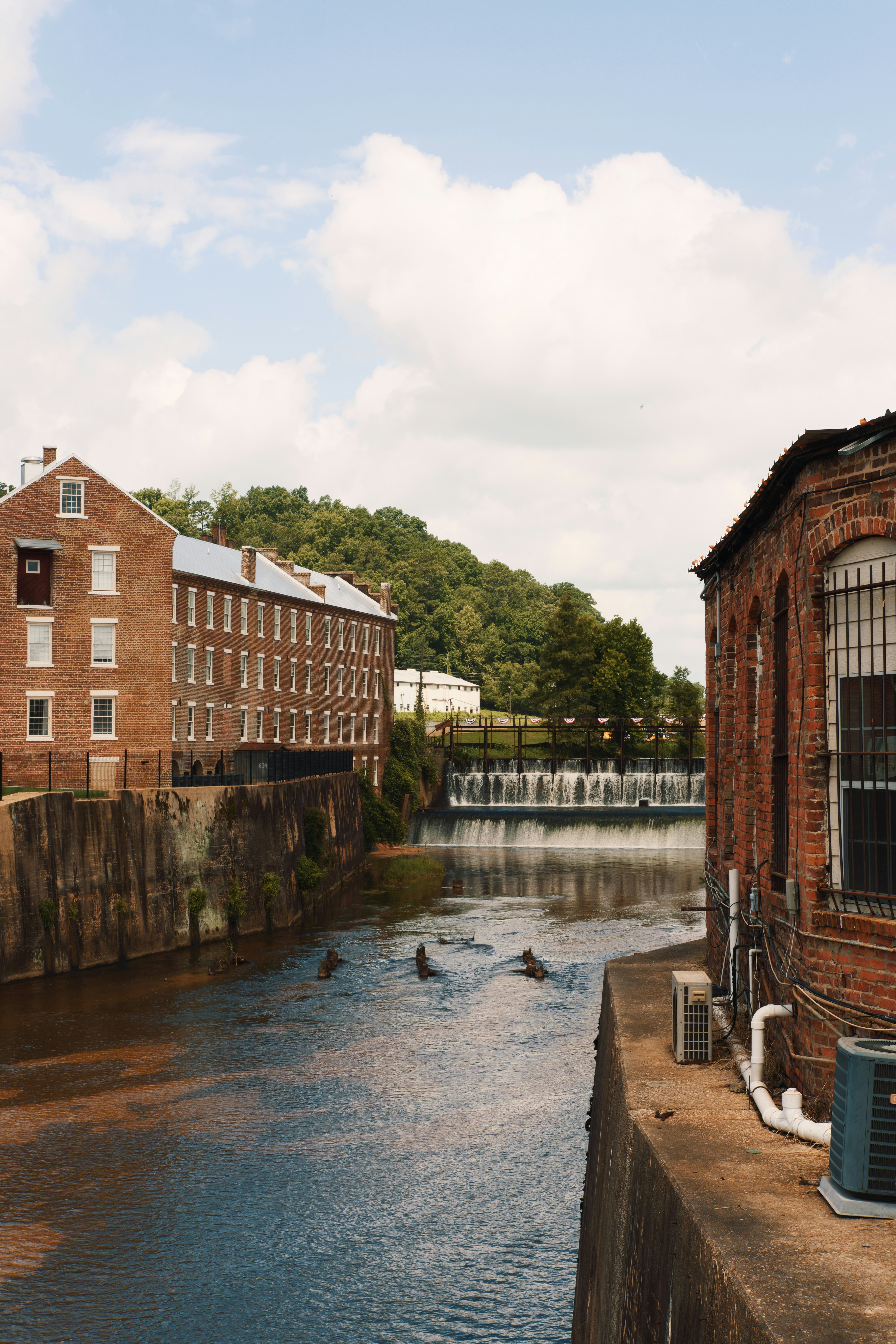 Brick buildings line a calm river, with a small waterfall and industrial structures along the water’s edge, suggesting a historic mill district setting.