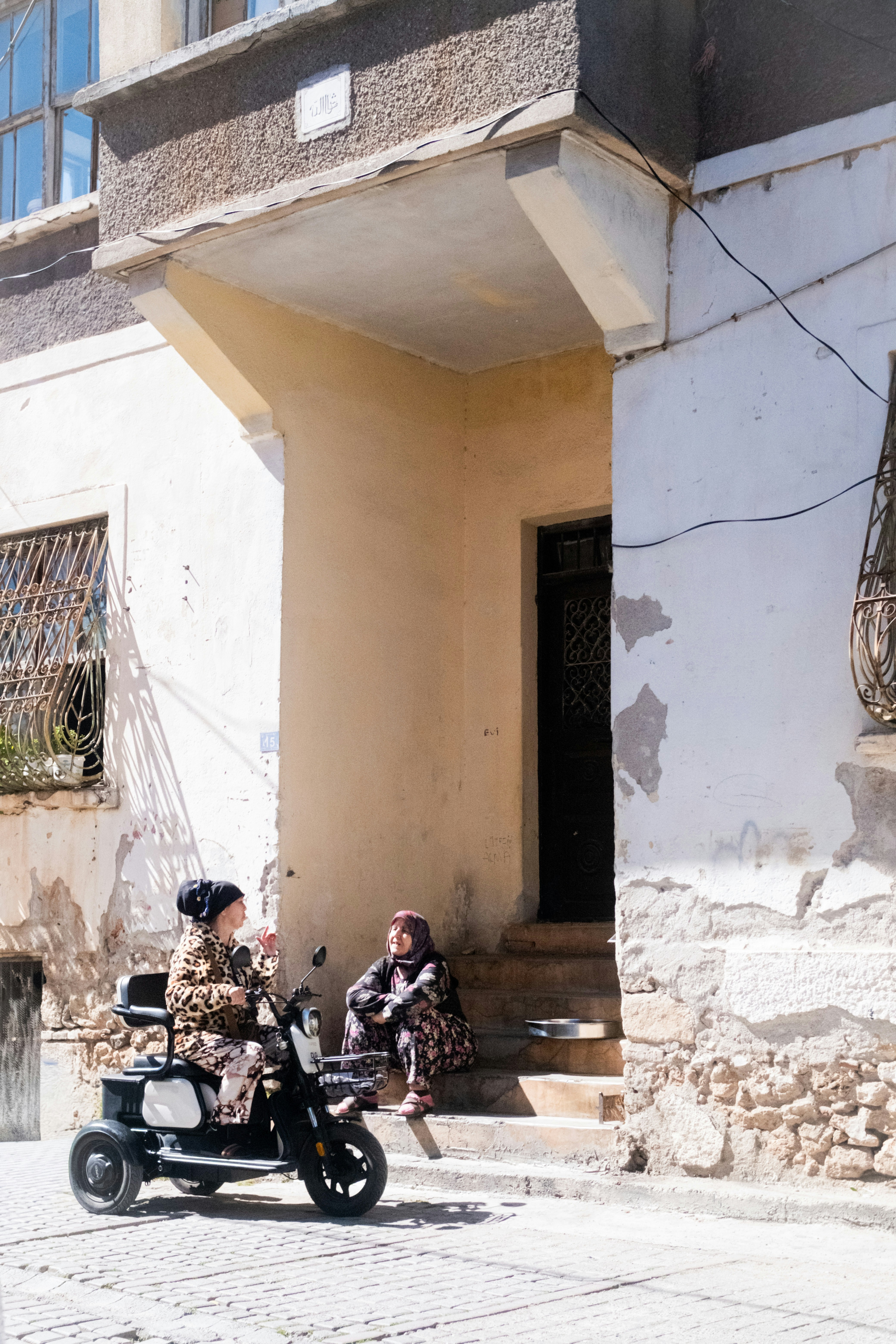 Two women sit and converse beside a retro scooter at the base of a weathered building; a doorway and a window frame the scene while warm light highlights their patterned clothing. This is a candid street photograph.