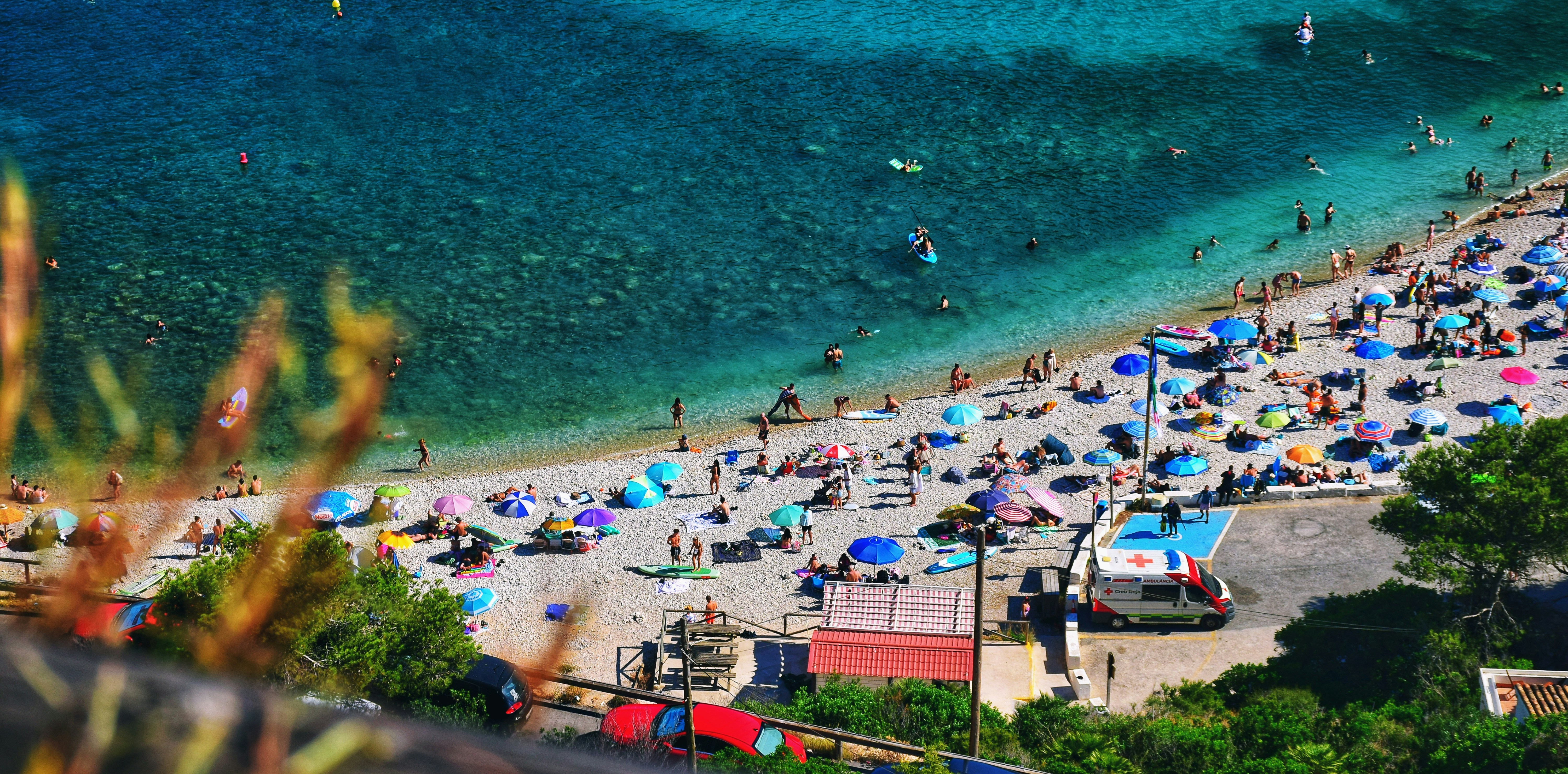Vibrant beach scene with sunbathers, umbrellas, and swimmers along a turquoise coastal shore.