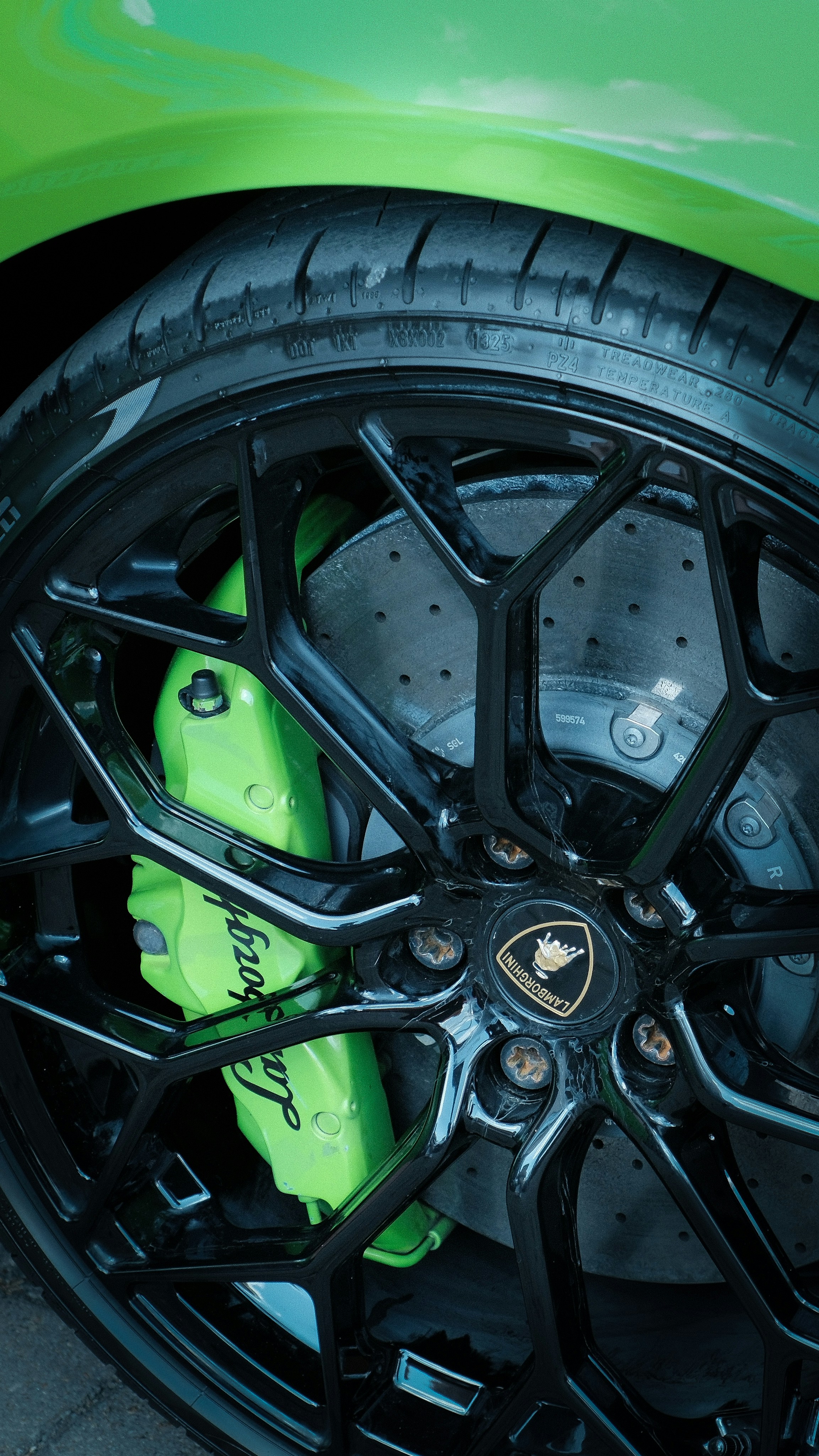 Close-up of a vibrant lime-green Lamborghini brake caliper behind a black alloy wheel, showcasing mechanical detail and perforated rotor within the brake assembly.