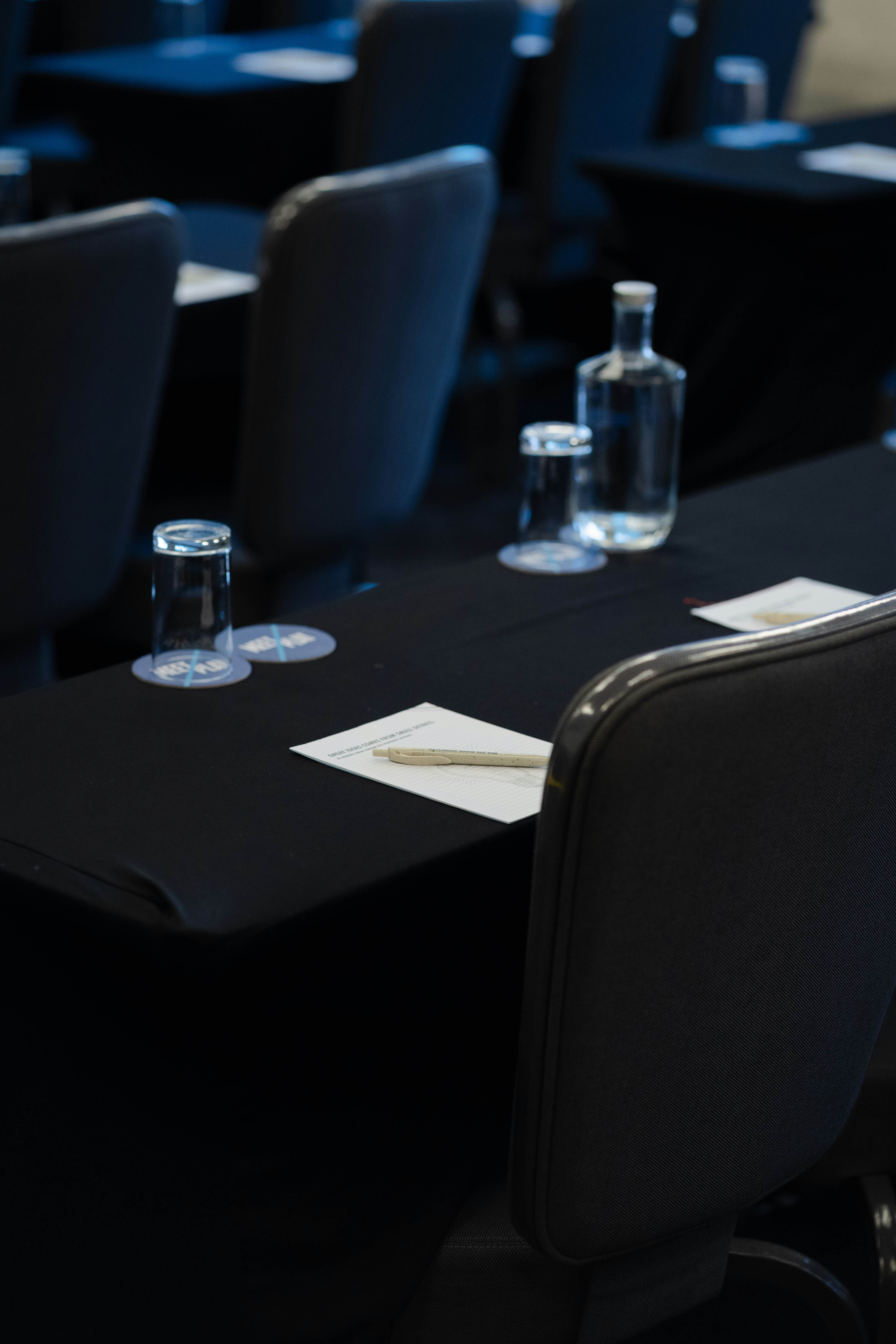 A dimly lit conference setup with neatly arranged black-cloth tables, glass water bottles, and pens on white notepads, conveying a formal meeting or presentation scene.
